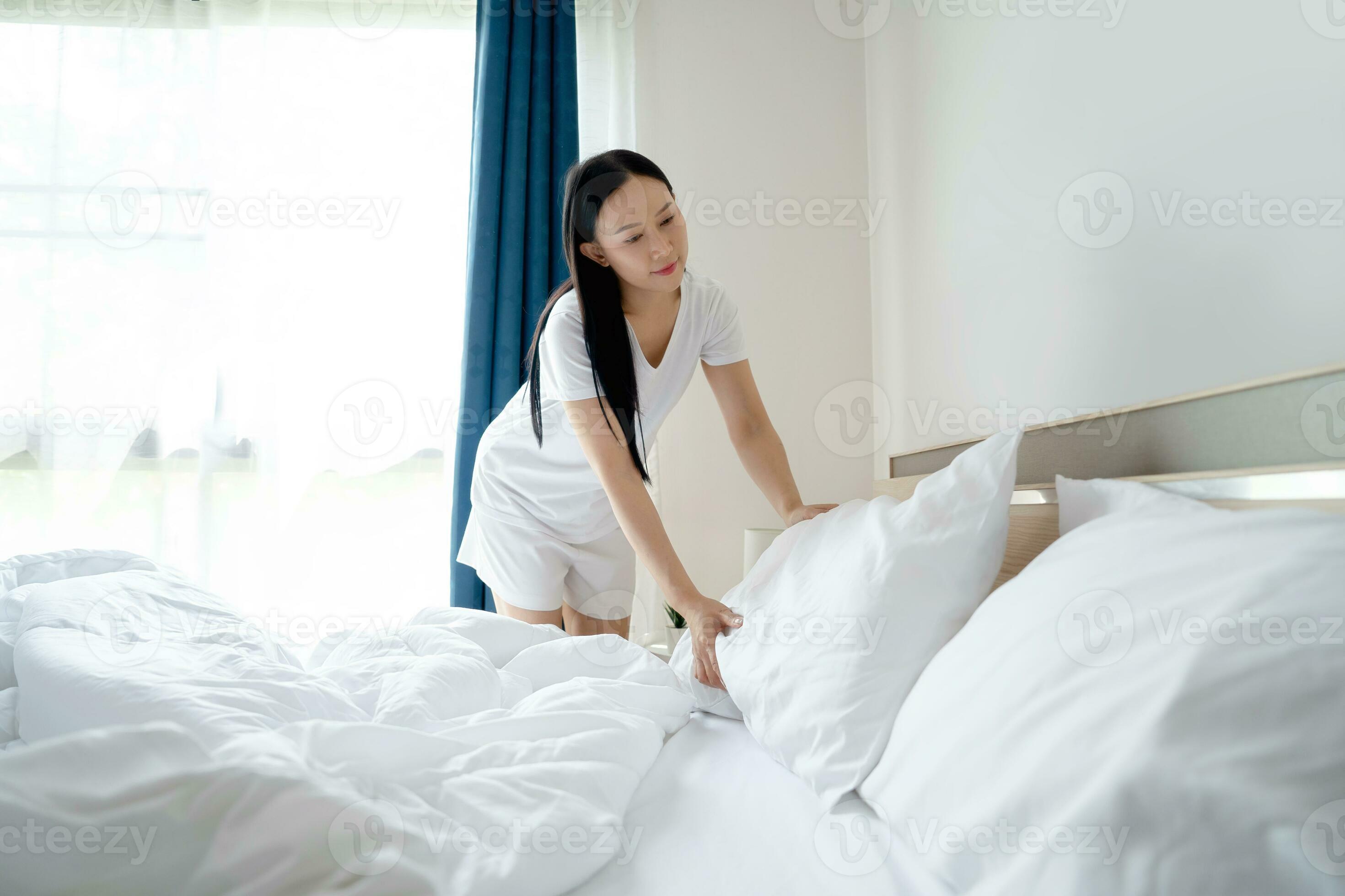 Asian woman with long black hair in white pajamas. She was making her bed in the morning after ...
