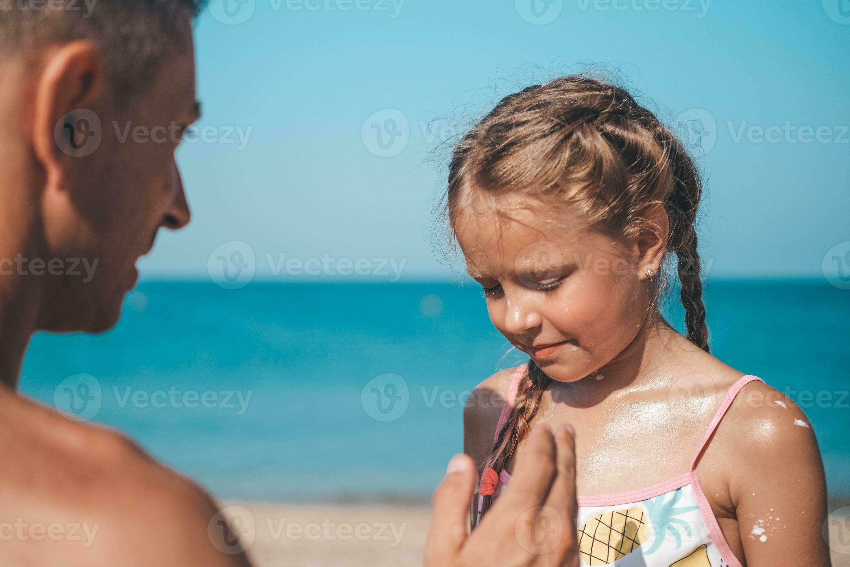 A father applies protective cream to his daughter's back at the beach. A man holds sunscreen ...