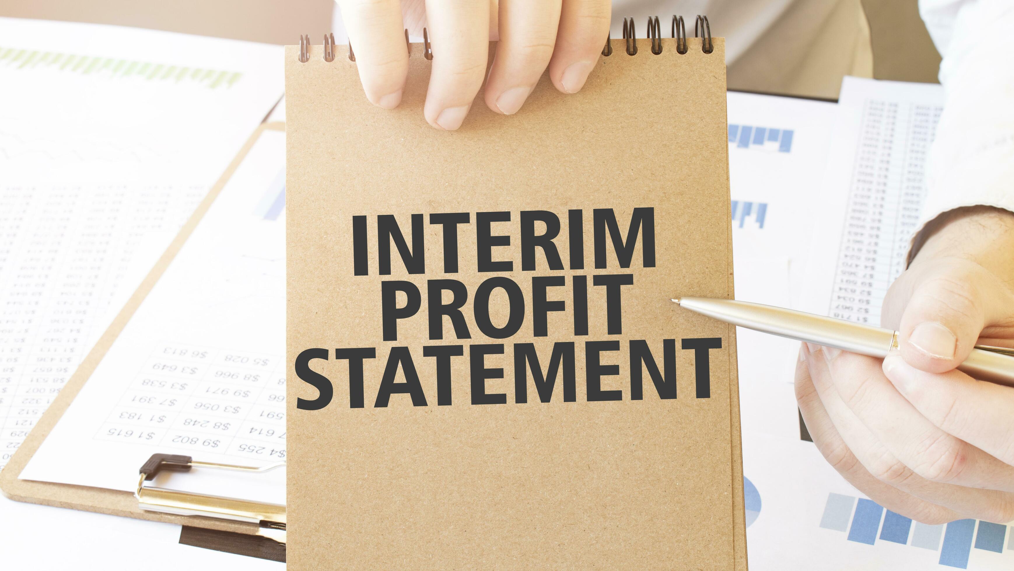 Text INTERIM PROFIT STATEMENT on brown paper notepad in businessman hands on the table with ...
