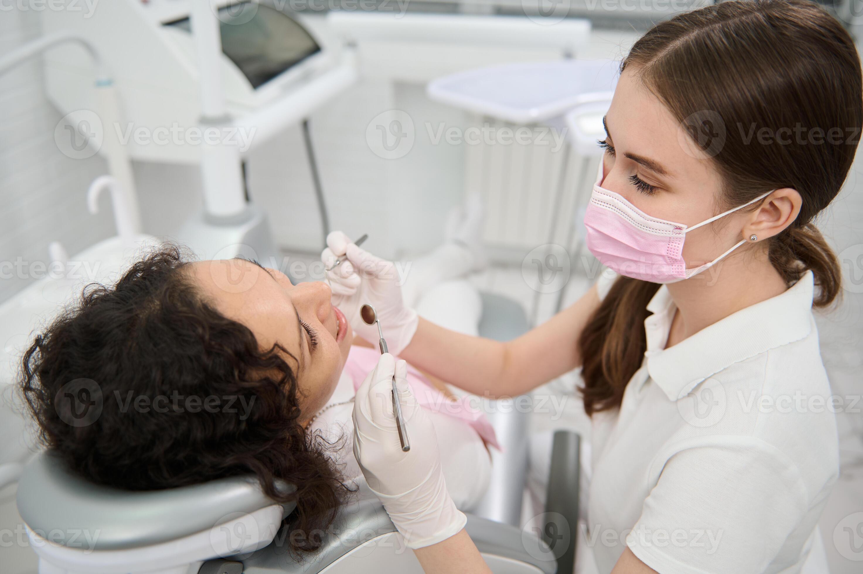 Dentist hygienist in medical protective mask examining the oral cavity of a female patient with ...