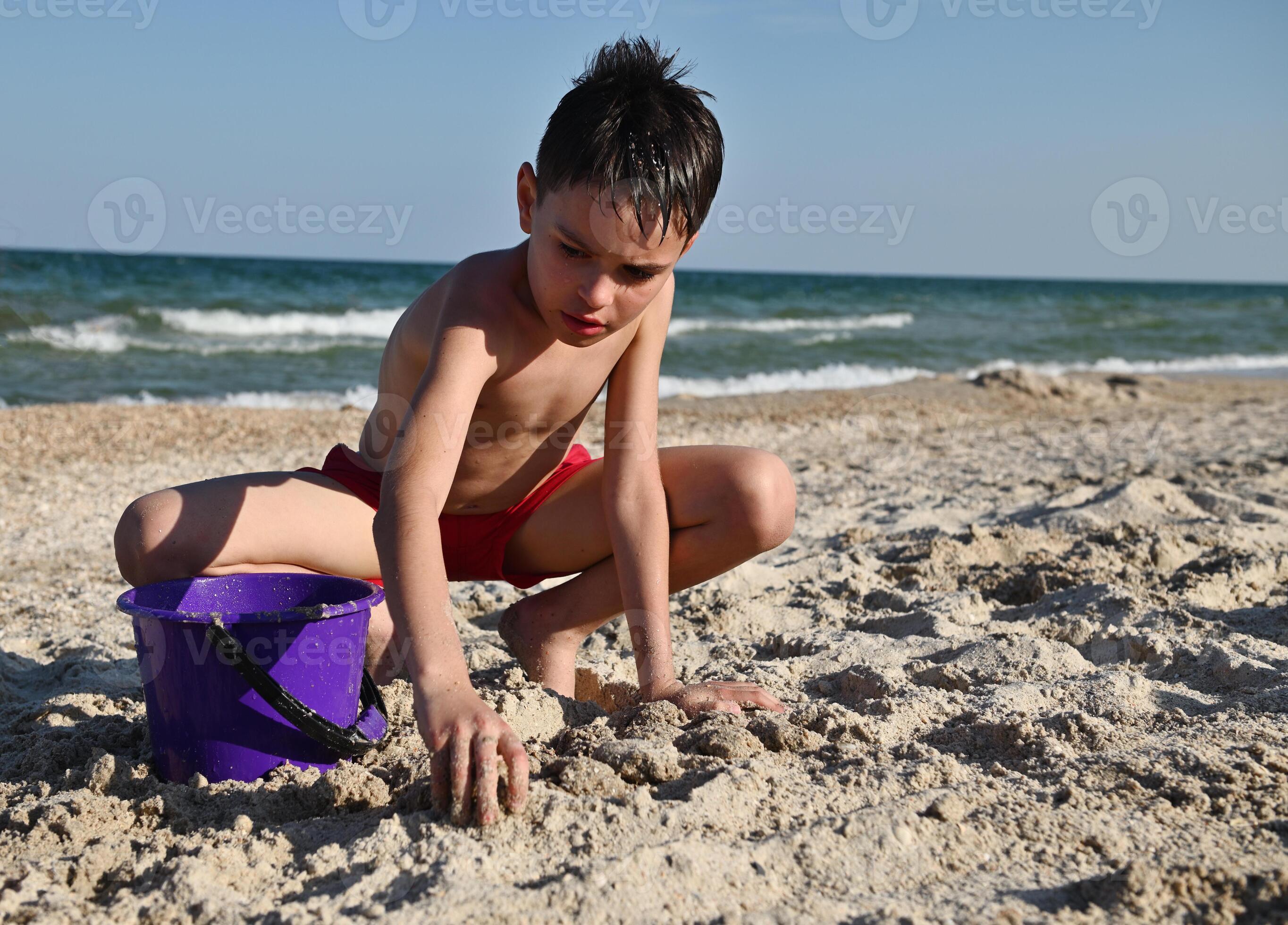 Cute teenage boy in red swimming trunks playing on the sandy beach. Adorable male kid builds ...