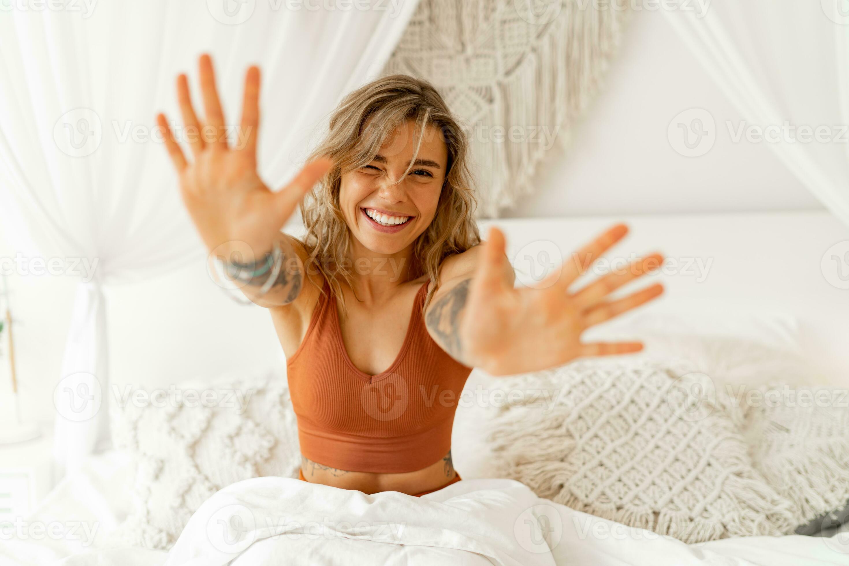 Smiling young woman stretching in bed waking up in cozy bedroom with bohemian interior style ...
