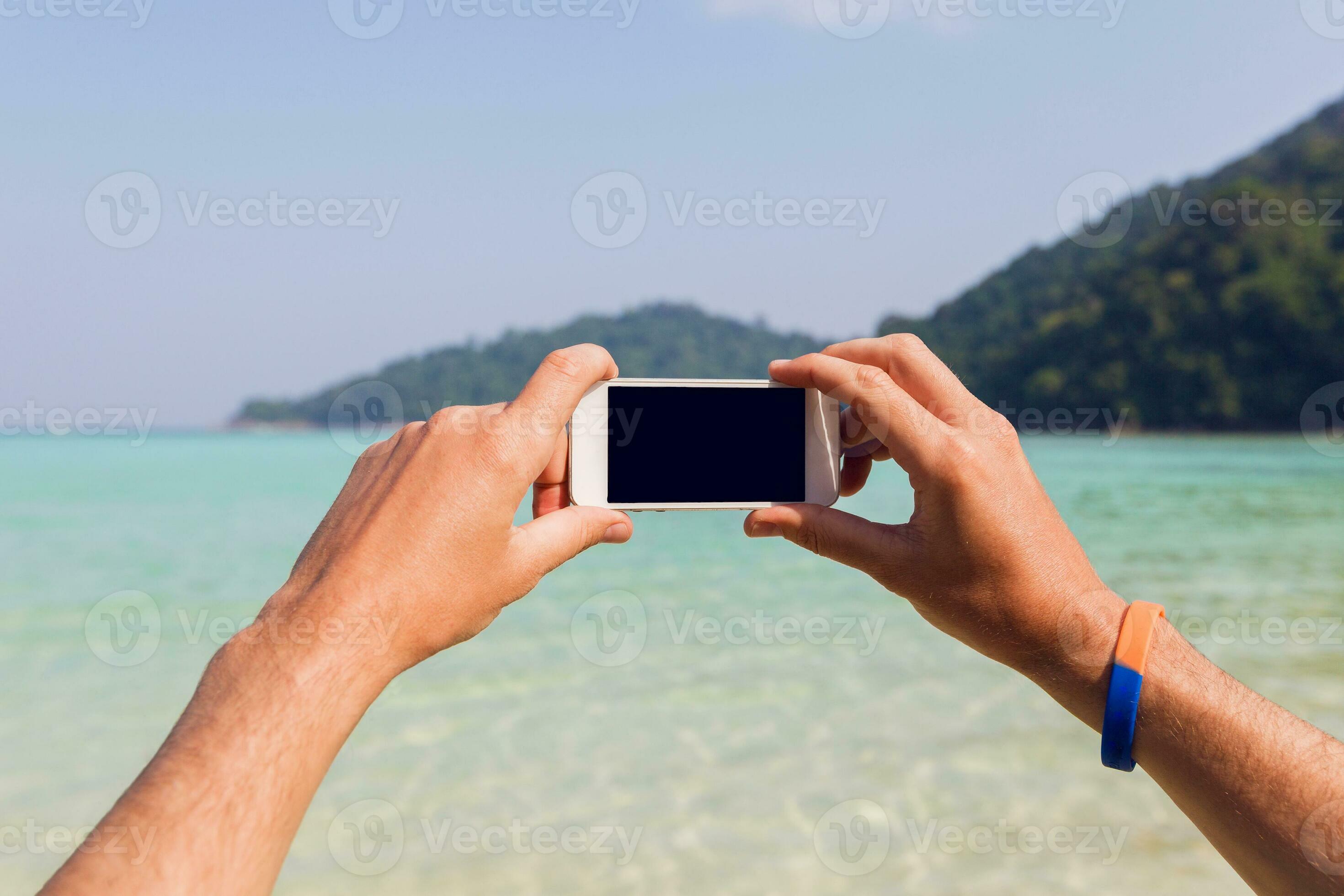 Man hands holding white mobile phone with black screen. Amazing blue sea and sky tropical island ...