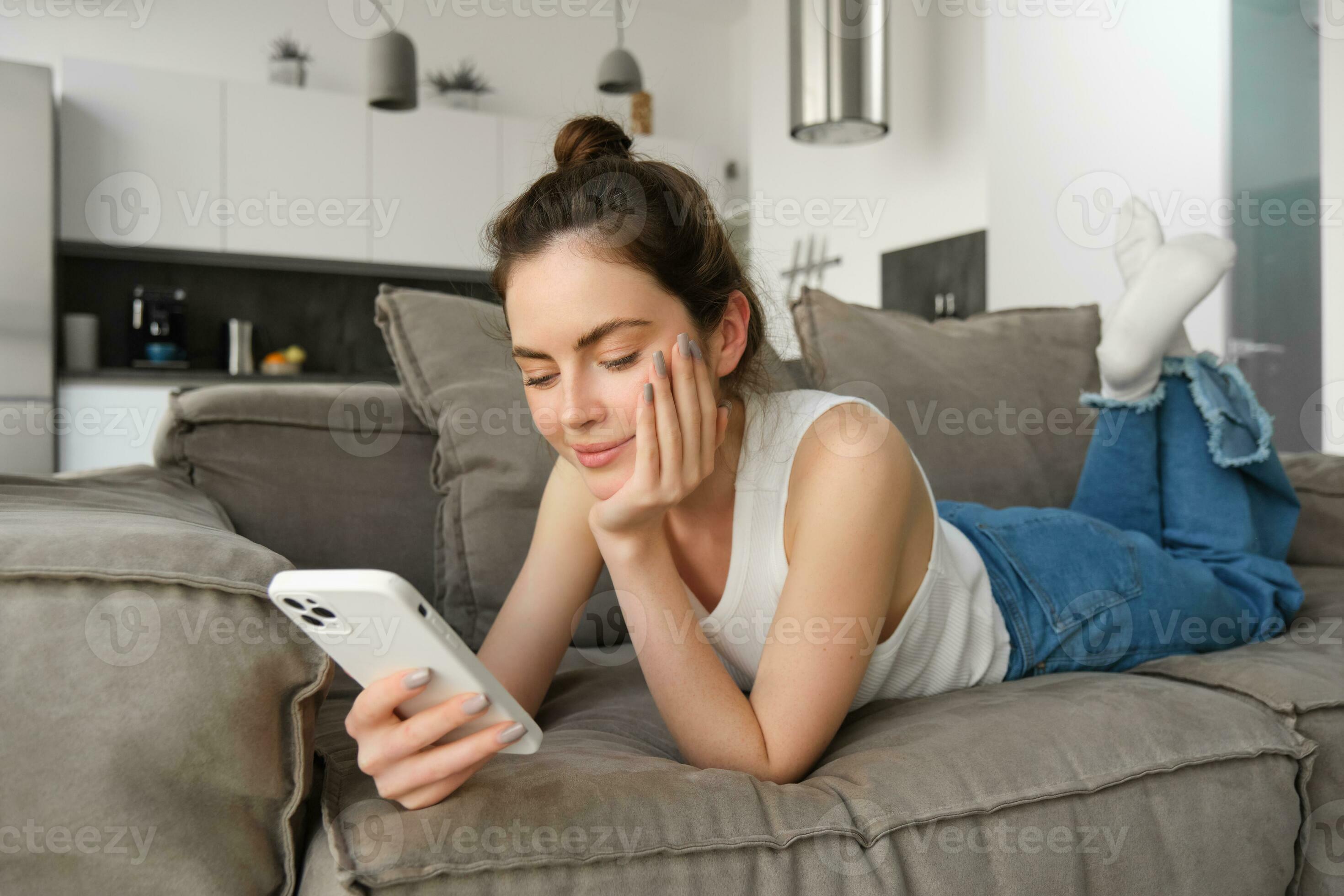 Portrait of cute young woman with smartphone, lying on couch and resting at home, scrolling ...