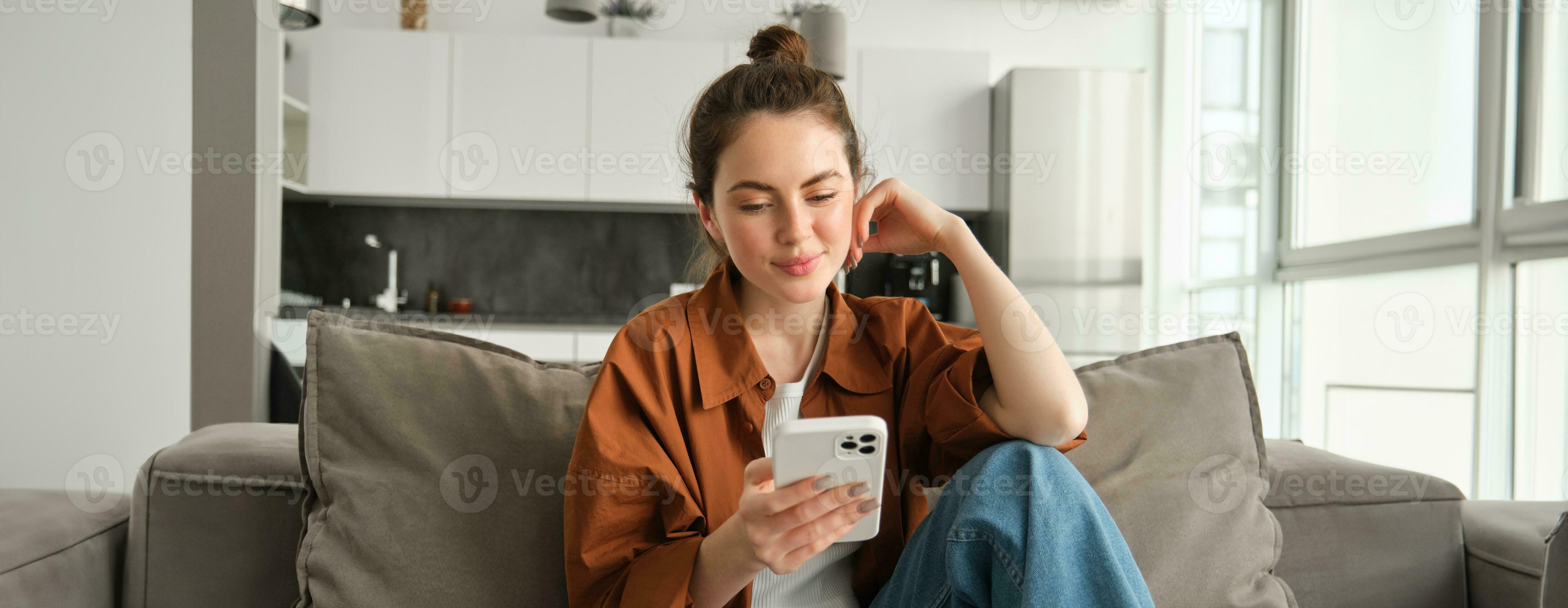 Portrait of happy young brunette woman sitting on sofa with smartphone, reading notification ...