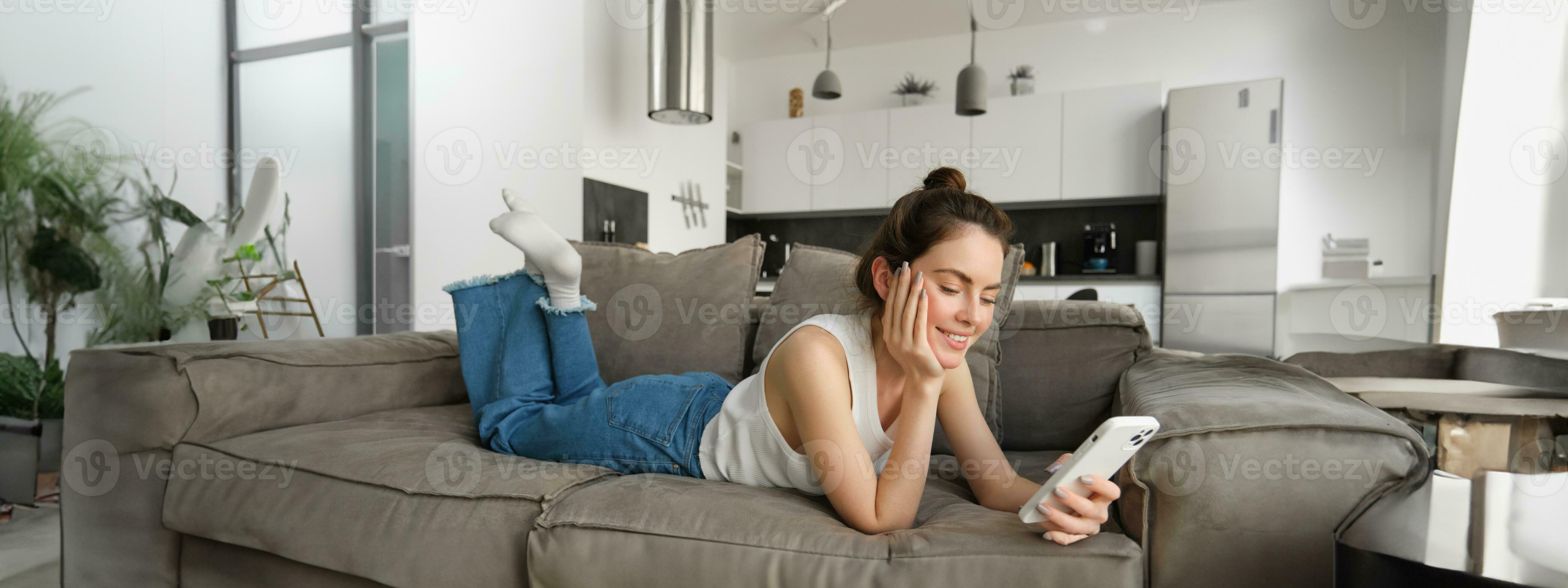 Portrait of cute young woman with smartphone, lying on couch and resting at home, scrolling ...