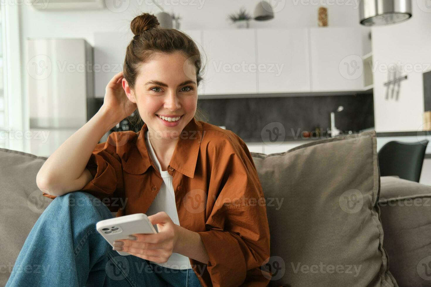 Beautiful smiling female model sitting on couch with smartphone, looking happy at camera ...