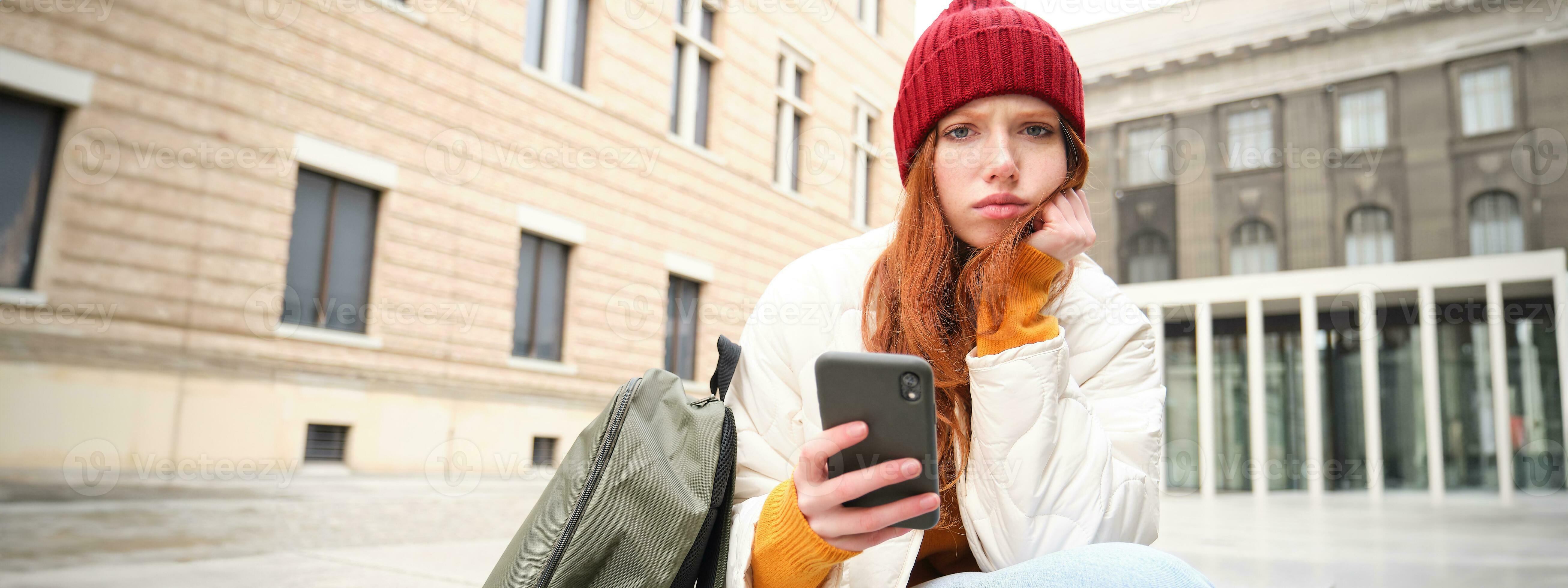 Redhead 20s years girl, sits with smartphone outside building, looks complicated and upset ...