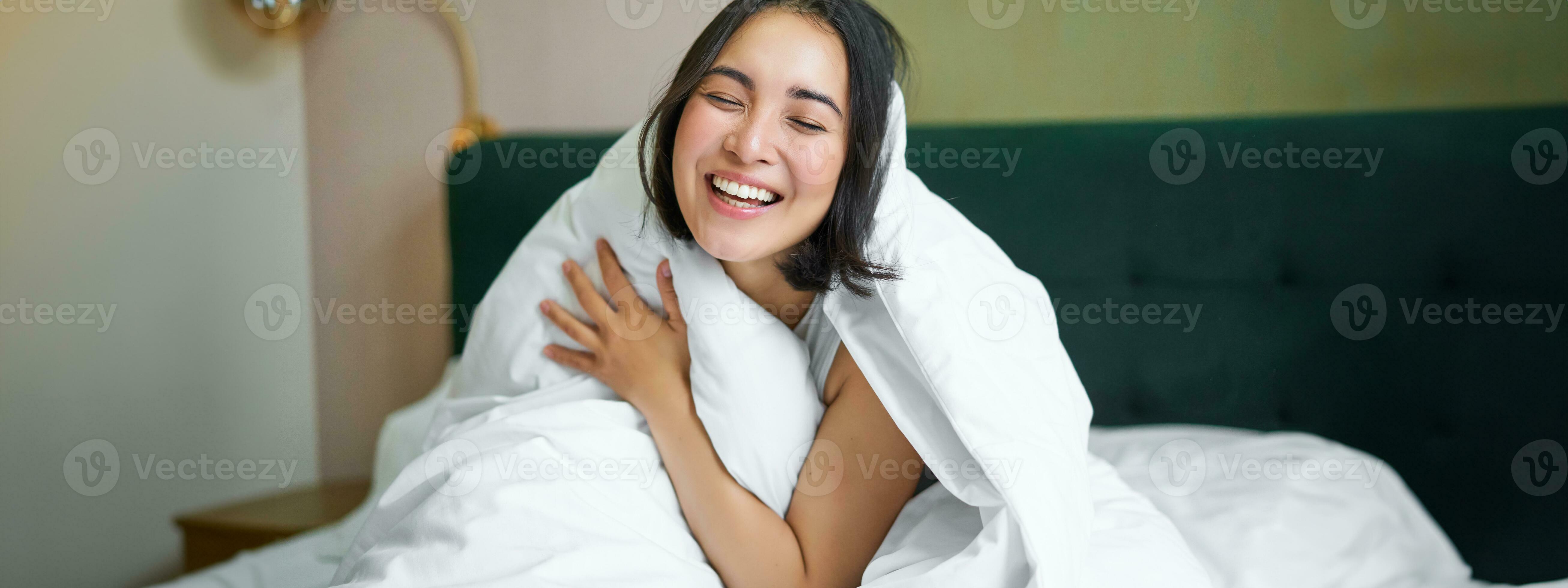 Beautiful Asian Woman Sitting On Bed Covered With White Duvet Smiling