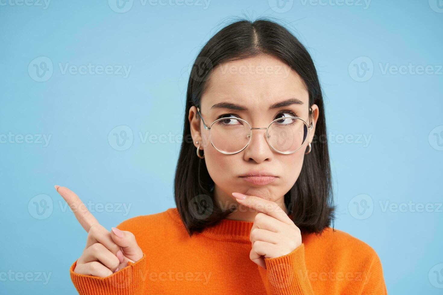 Image Of Thoughtful Asian Woman Frowns Wears Glasses Points Left And Thinks Makes Serious