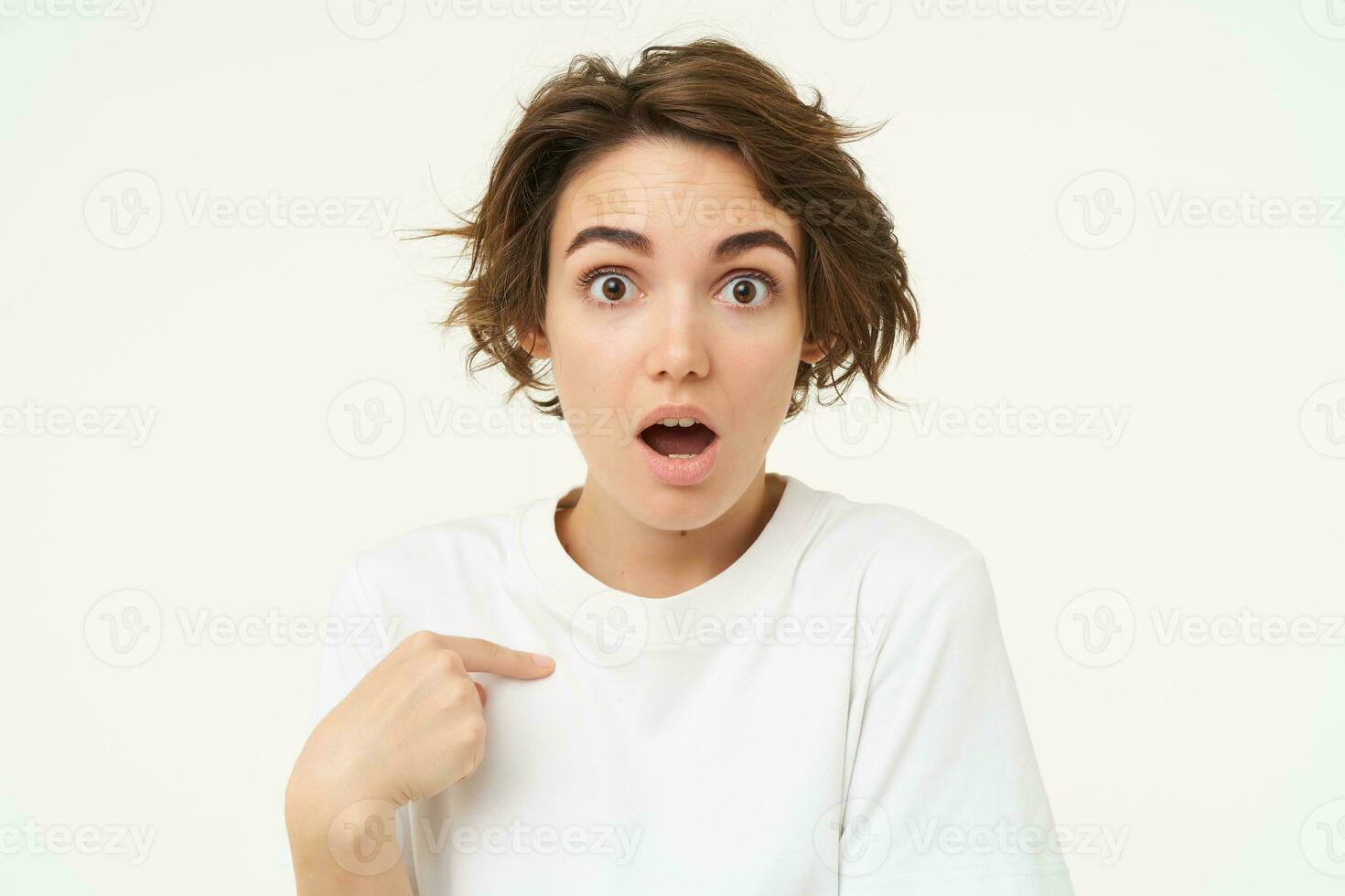 Portrait of girl with surprised face, points at herself amazed, stands isolated over white ...
