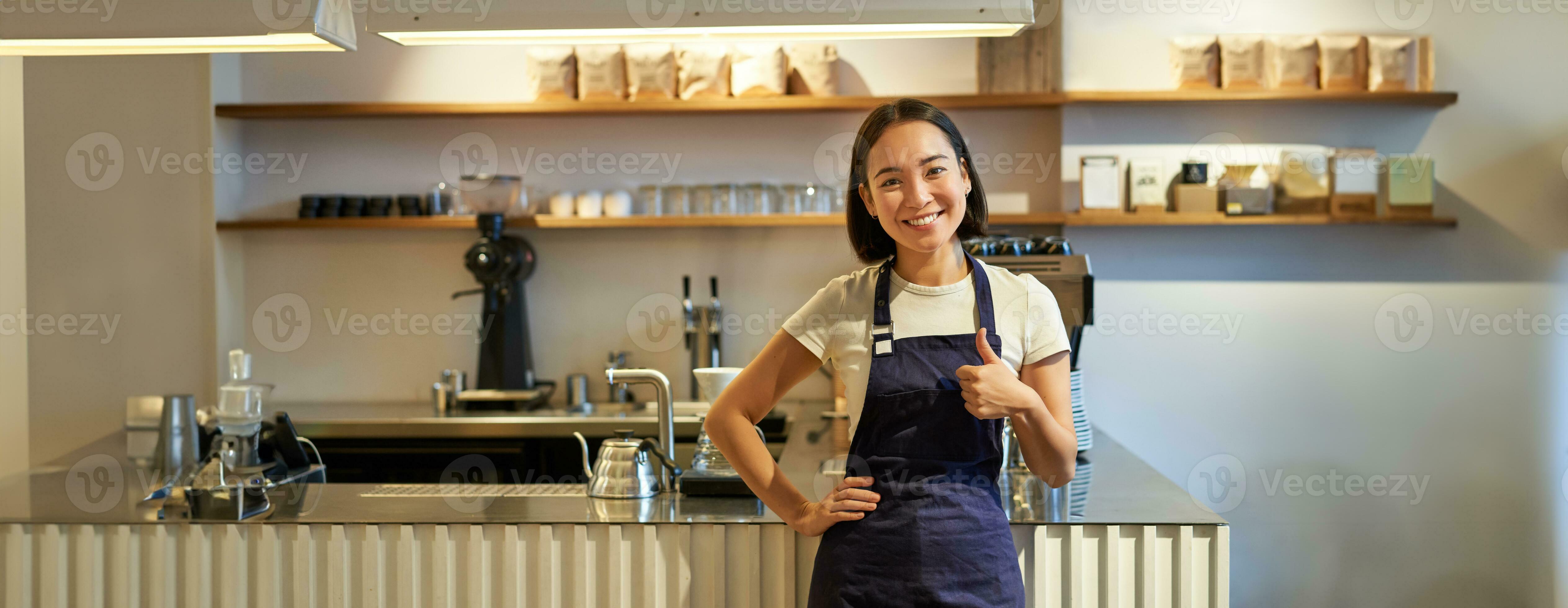 Smiling girl student working part-time in cafe, barista shows thumbs up, wears apron, stands ...