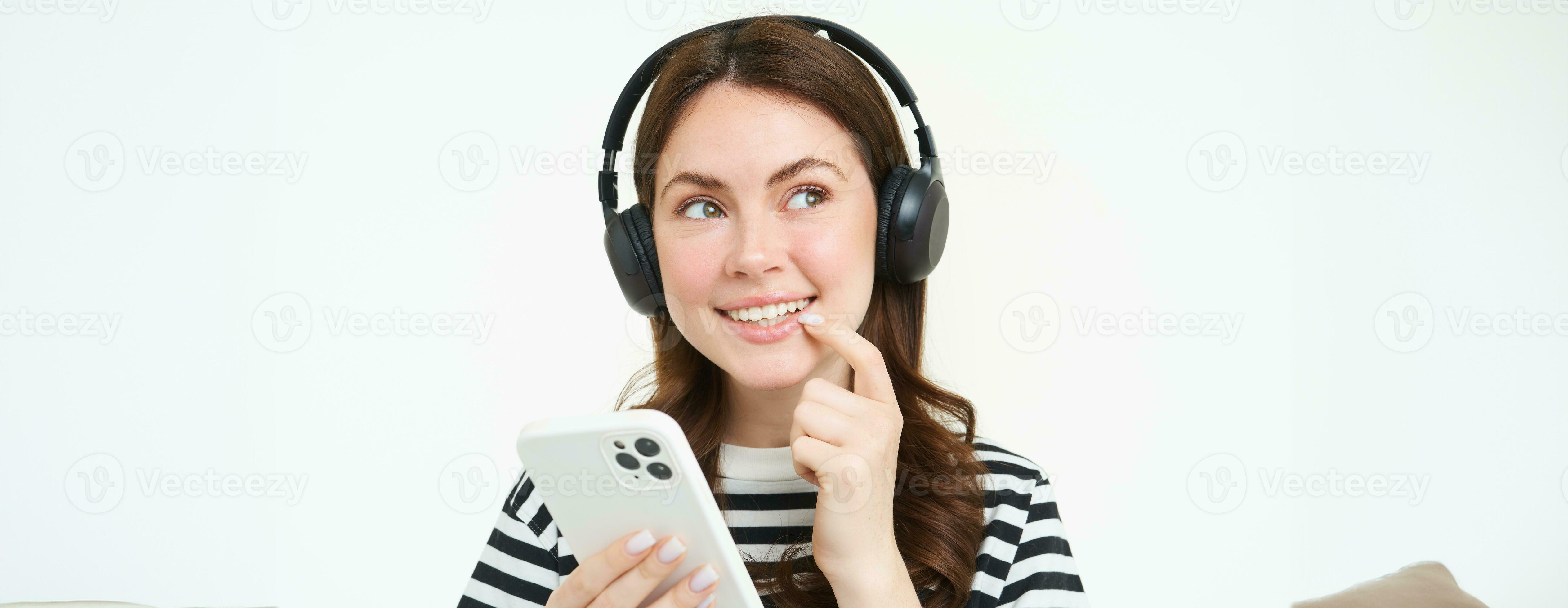 Image of girl in headphones, smiling while thinking, making decision, holding smartphone, making ...