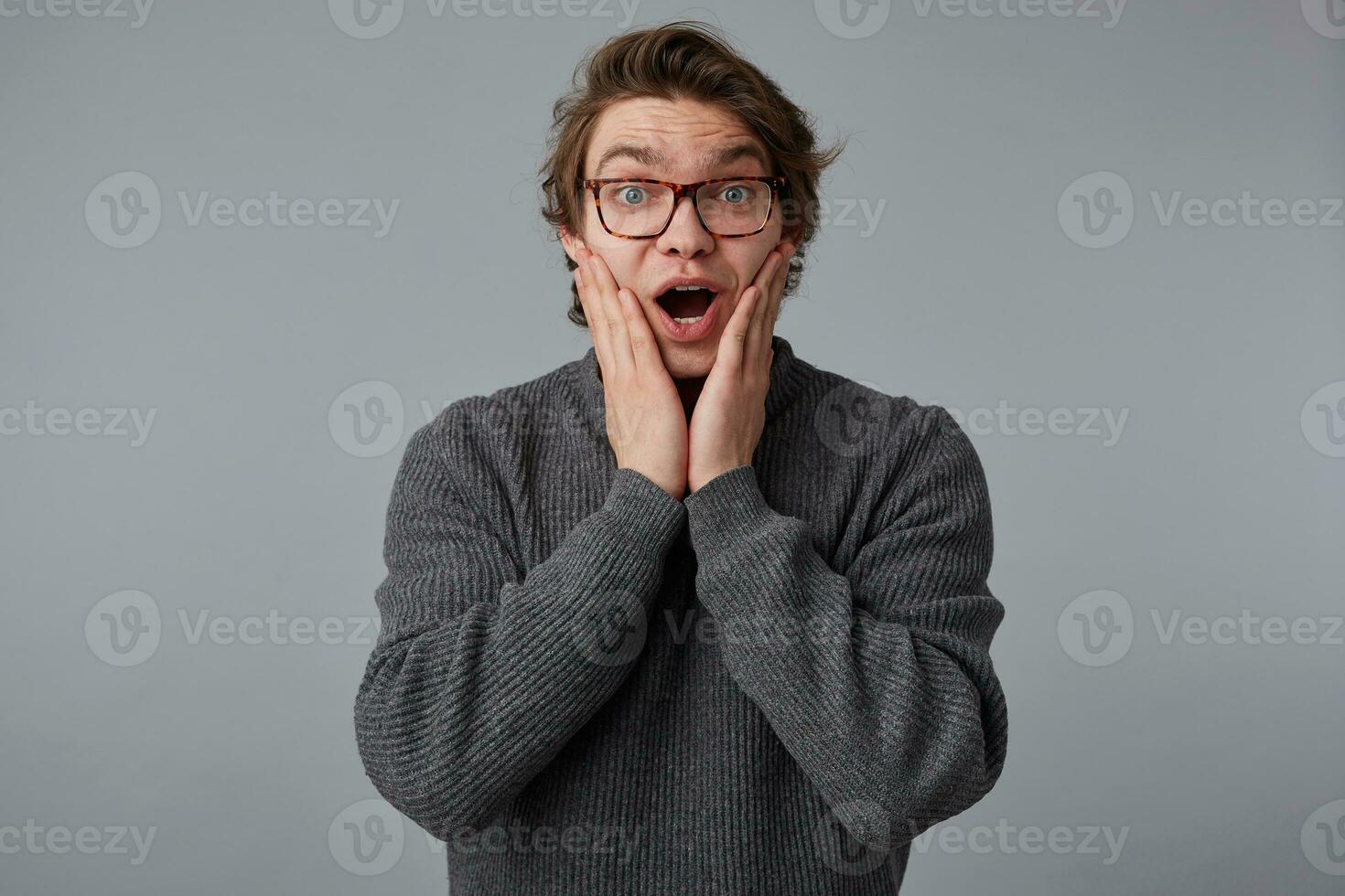 Young Shocked Guy With Glasses Wears In Gray Sweater Stands Over Gray