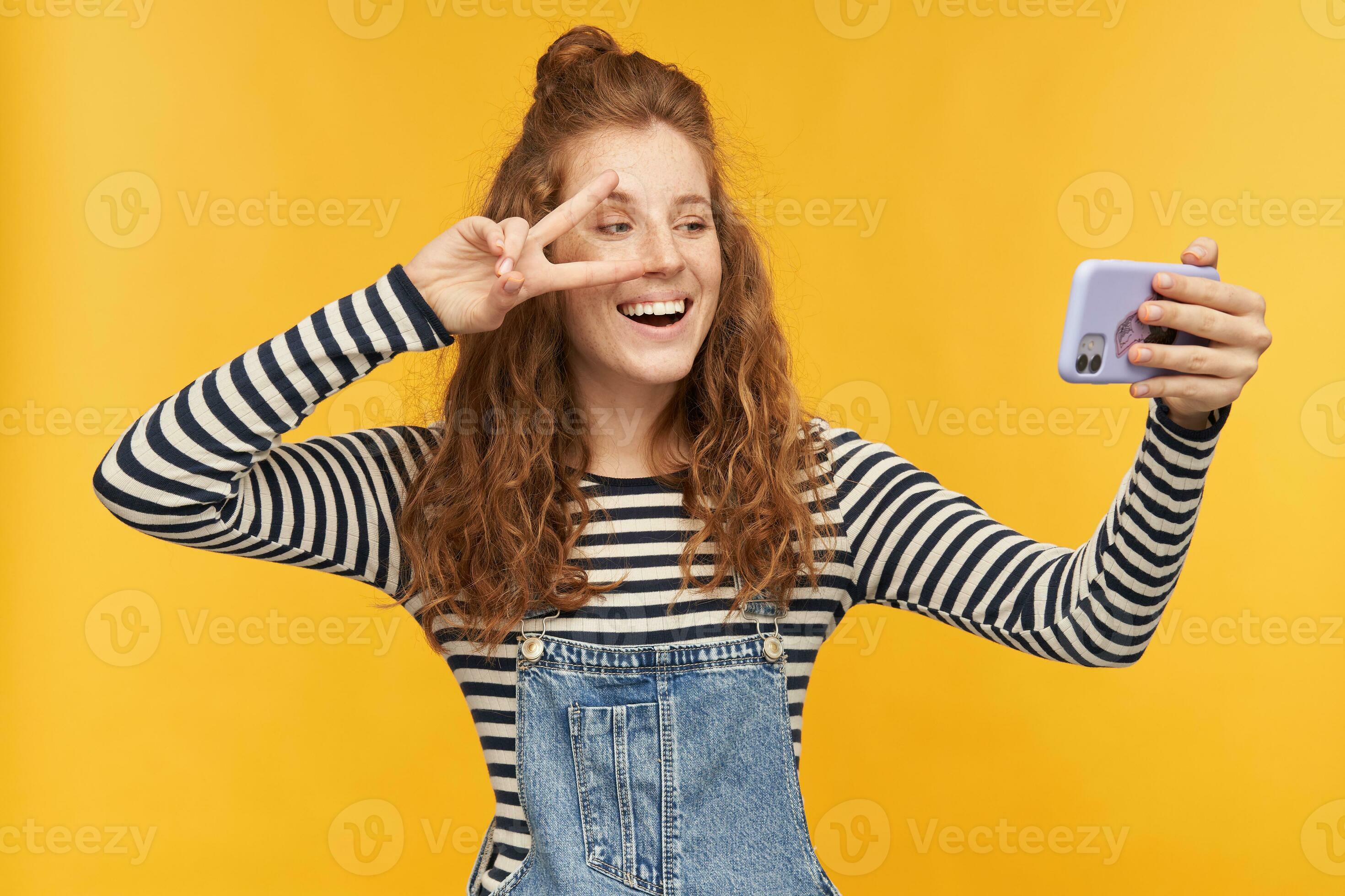 Indoor Portrait Of Young Happy Female Wears Stripped Shirt And Denim Overalls Shows V Sign