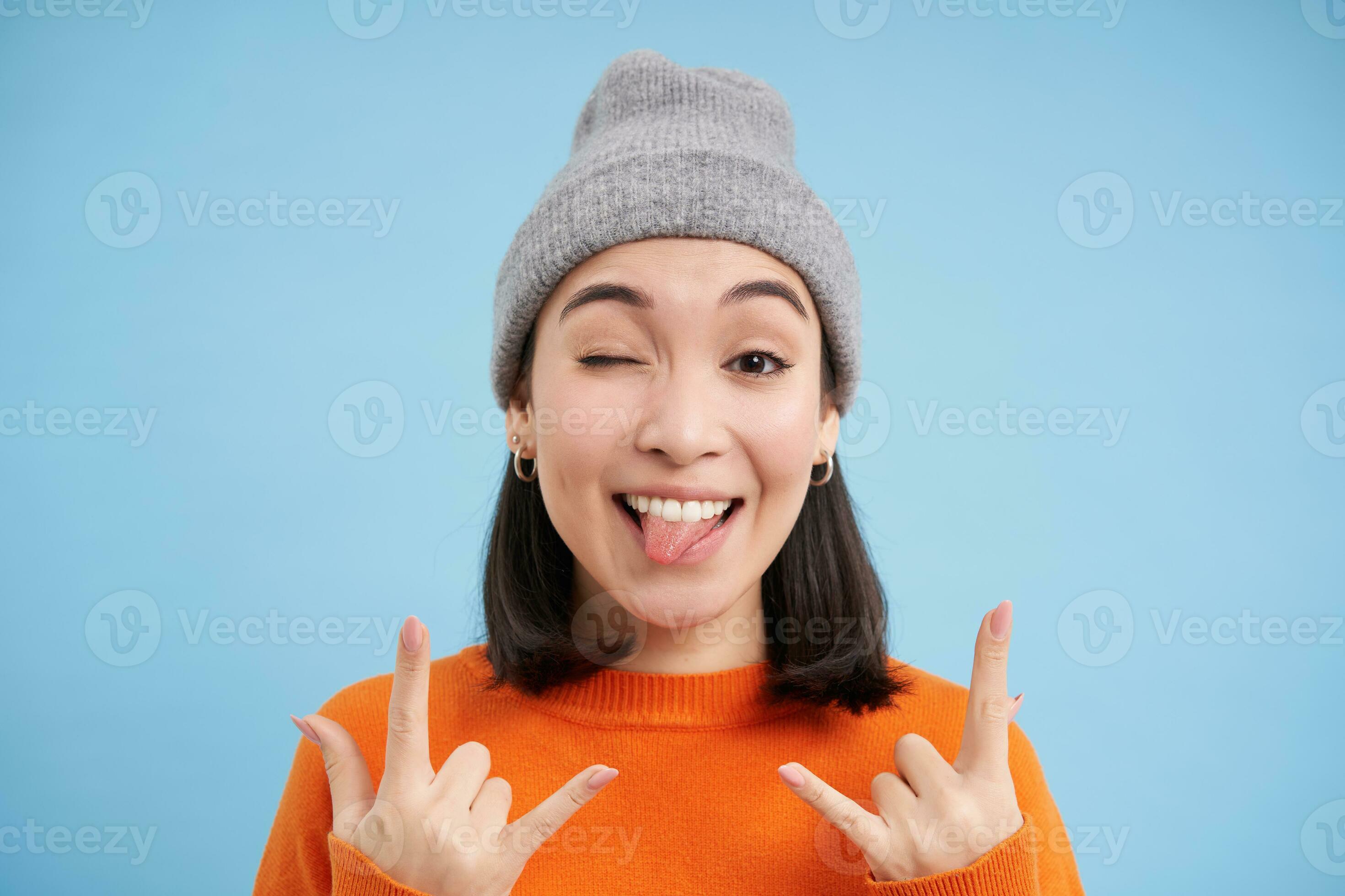 Close up portrait of funny and carefree asian girl in beanie, shows rock n roll, heavy metal ...