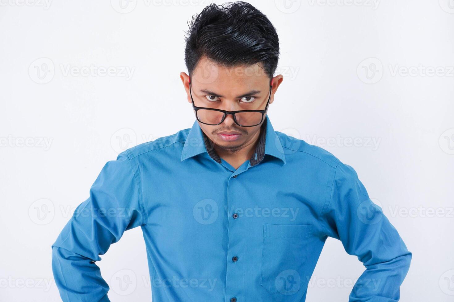 angry asian man with glasses standing with hands on waist and looking at camera wearing blue ...