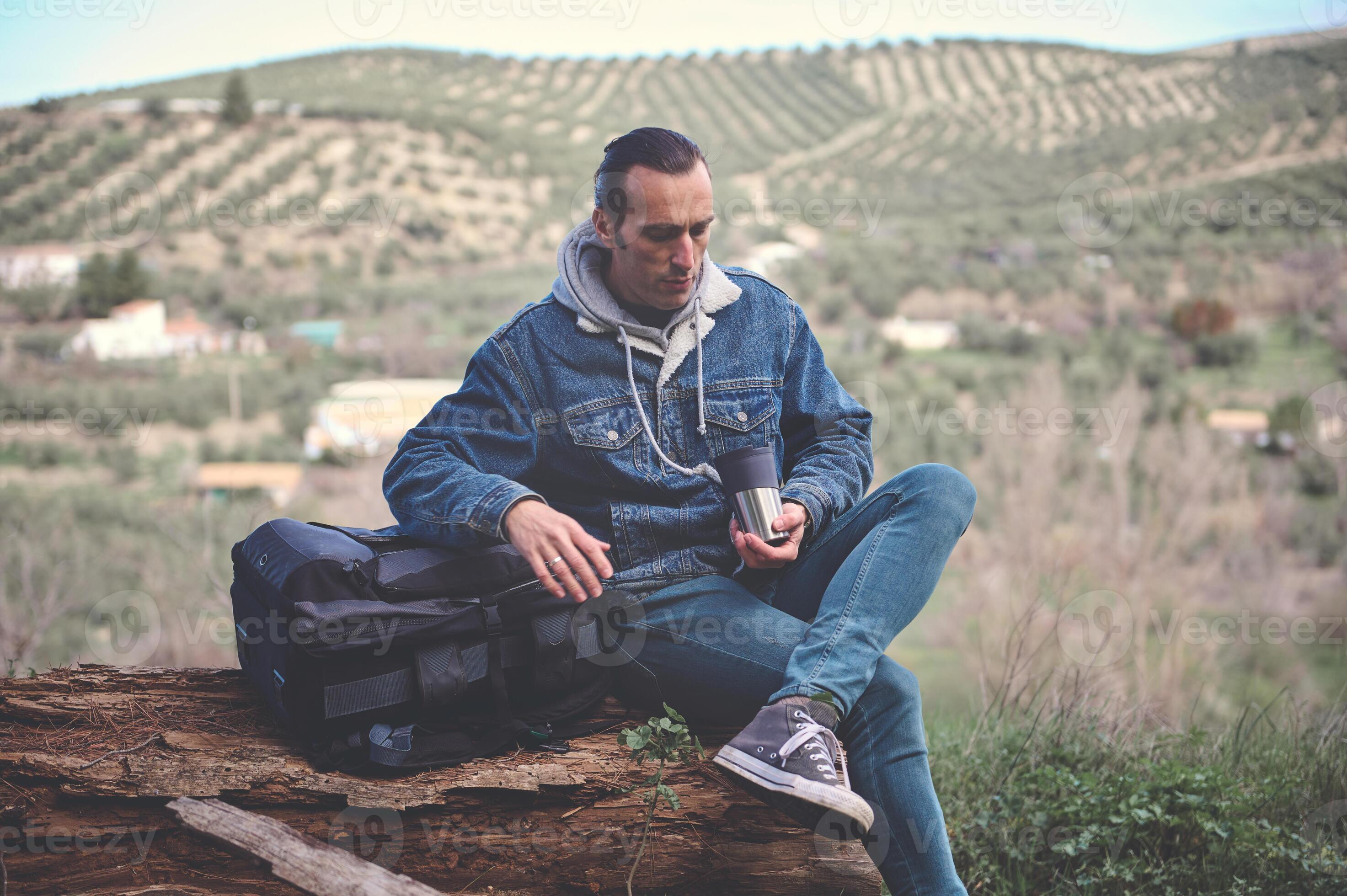 Backpacked adventurer traveler man, sitting on log in forest against mountains, drinking coffee ...