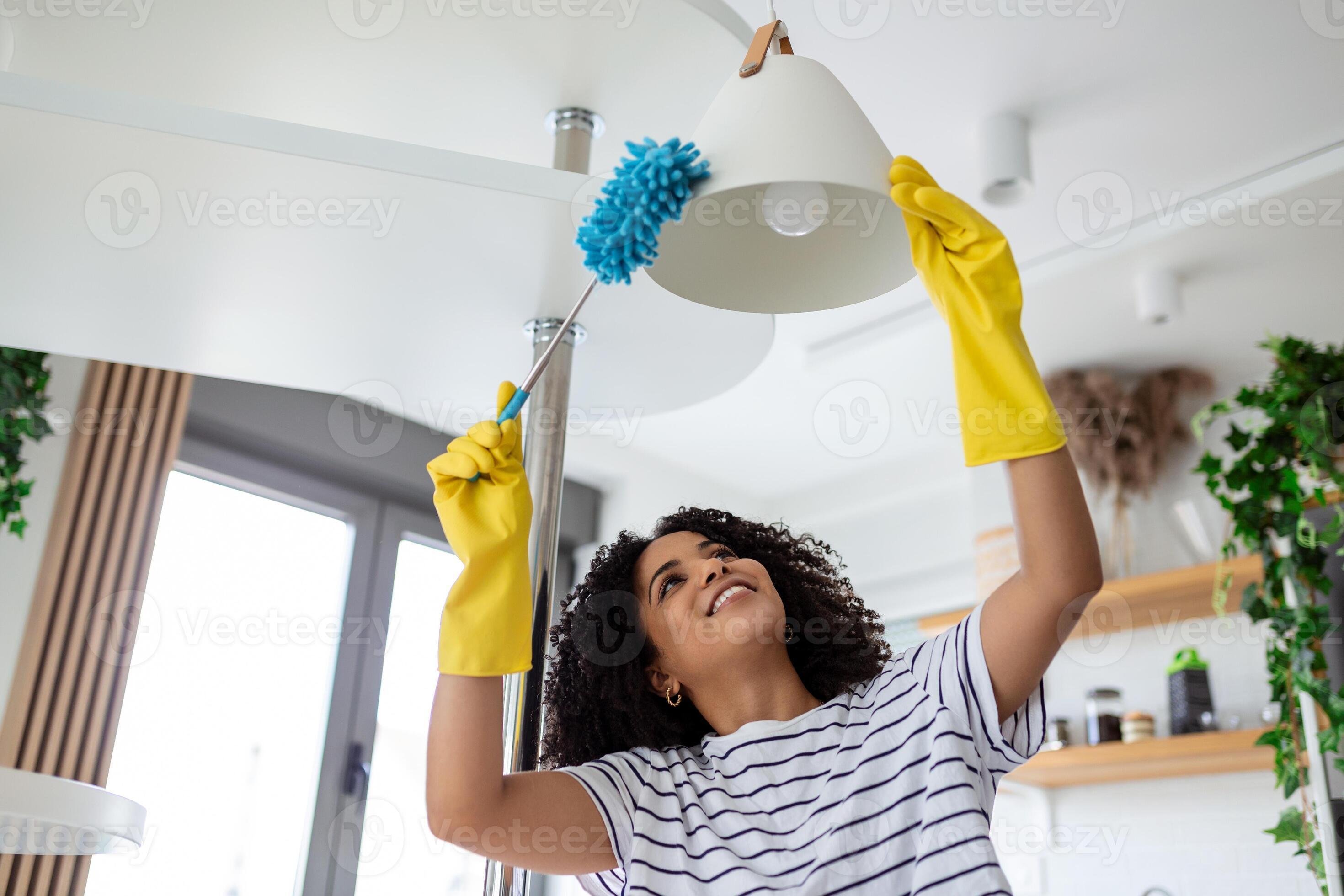 Young housekeeper in apron dusting the chandelier by duster carefully cleaning in living room at ...