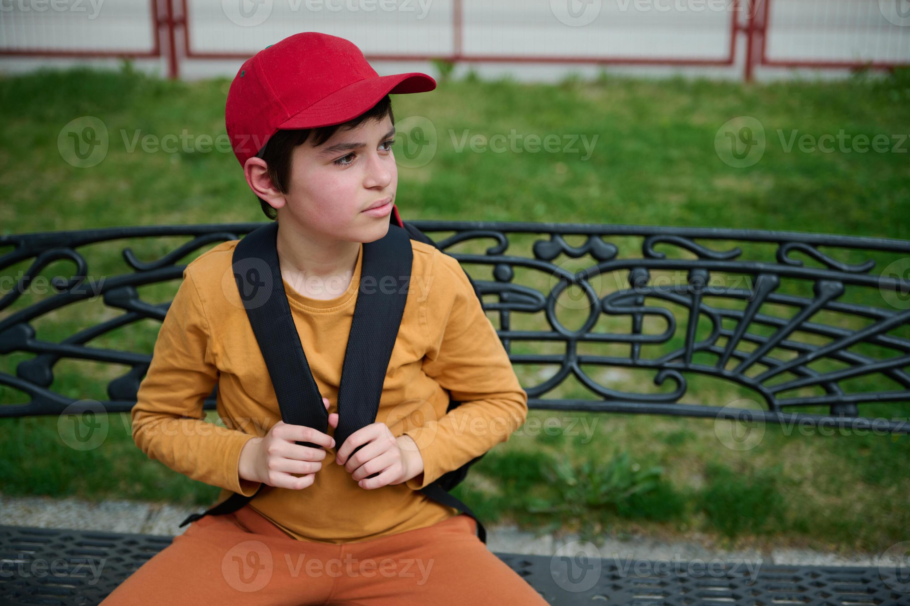 Backpacked Middle Eastern teenager, schoolboy looking aside, sitting on a bench in the park ...