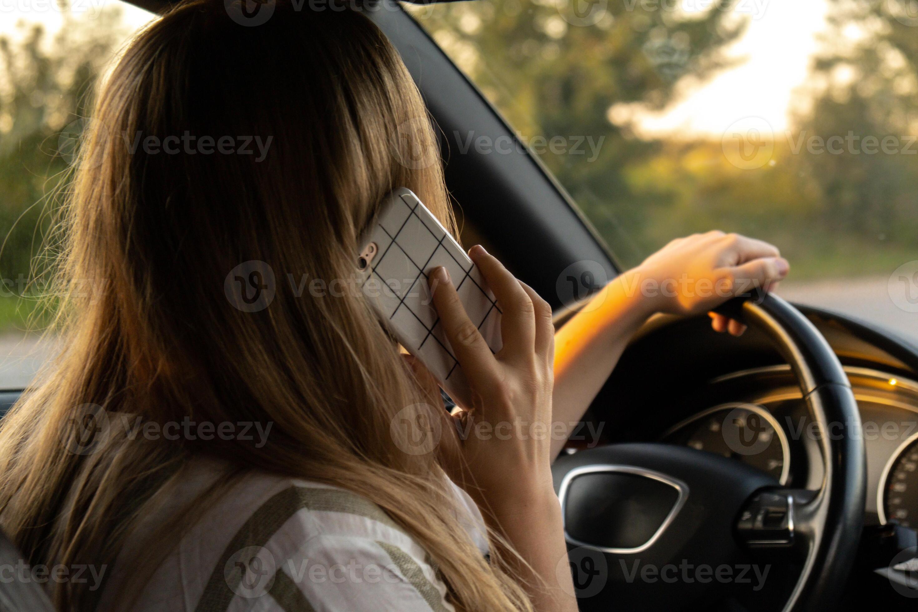 Young woman using mobile phone while driving car on highway road during sunset. Womandriver has ...