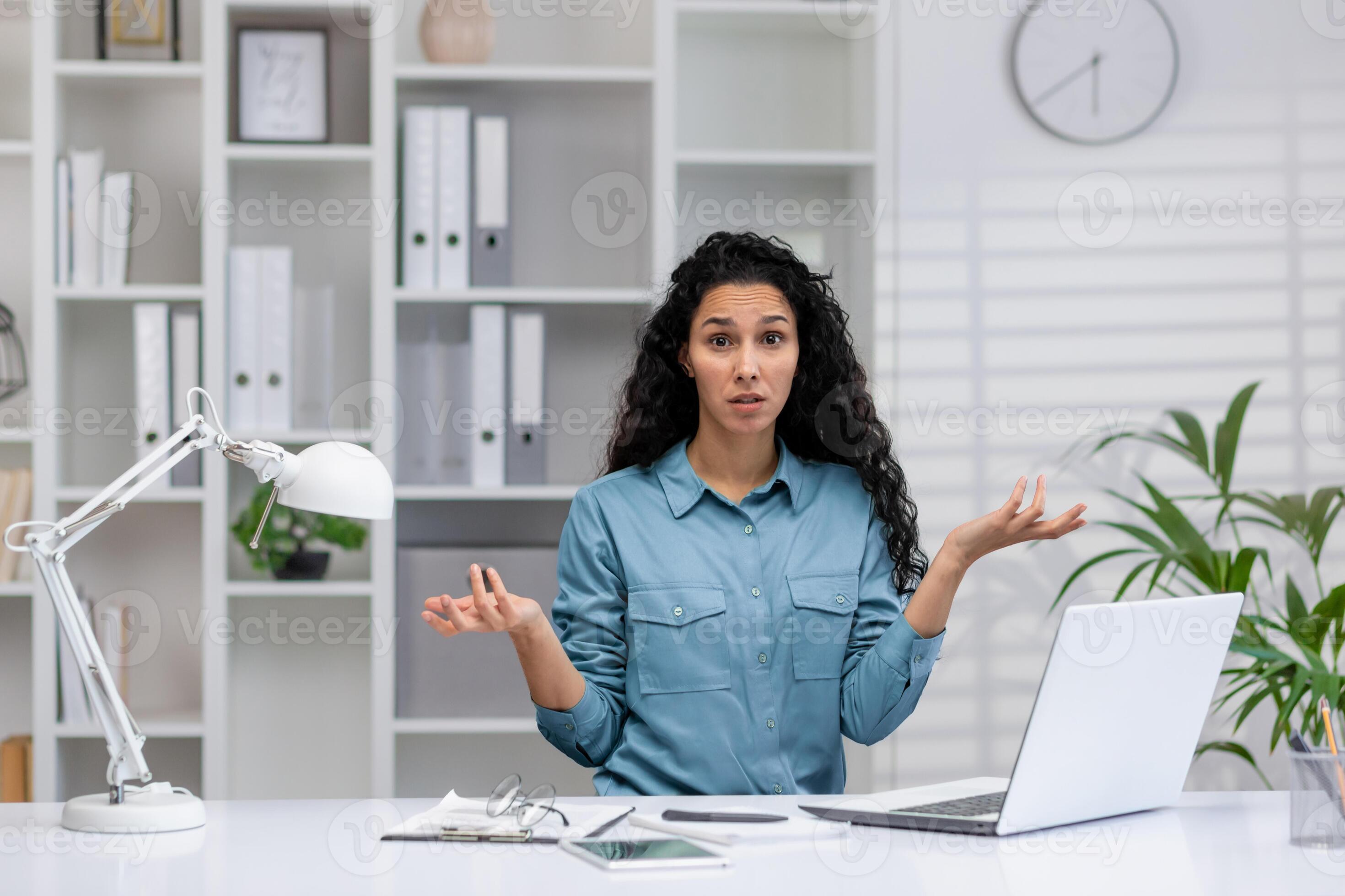 Perplexed Hispanic woman at her home office desk expressing confusion while using a laptop ...