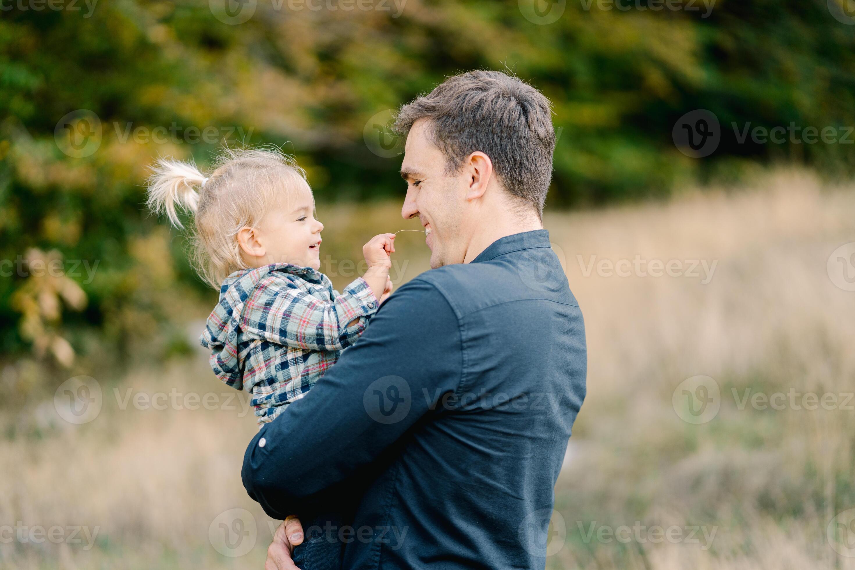 Little girl touches a blade of grass to the face of a smiling dad, sitting in his arms. Side ...