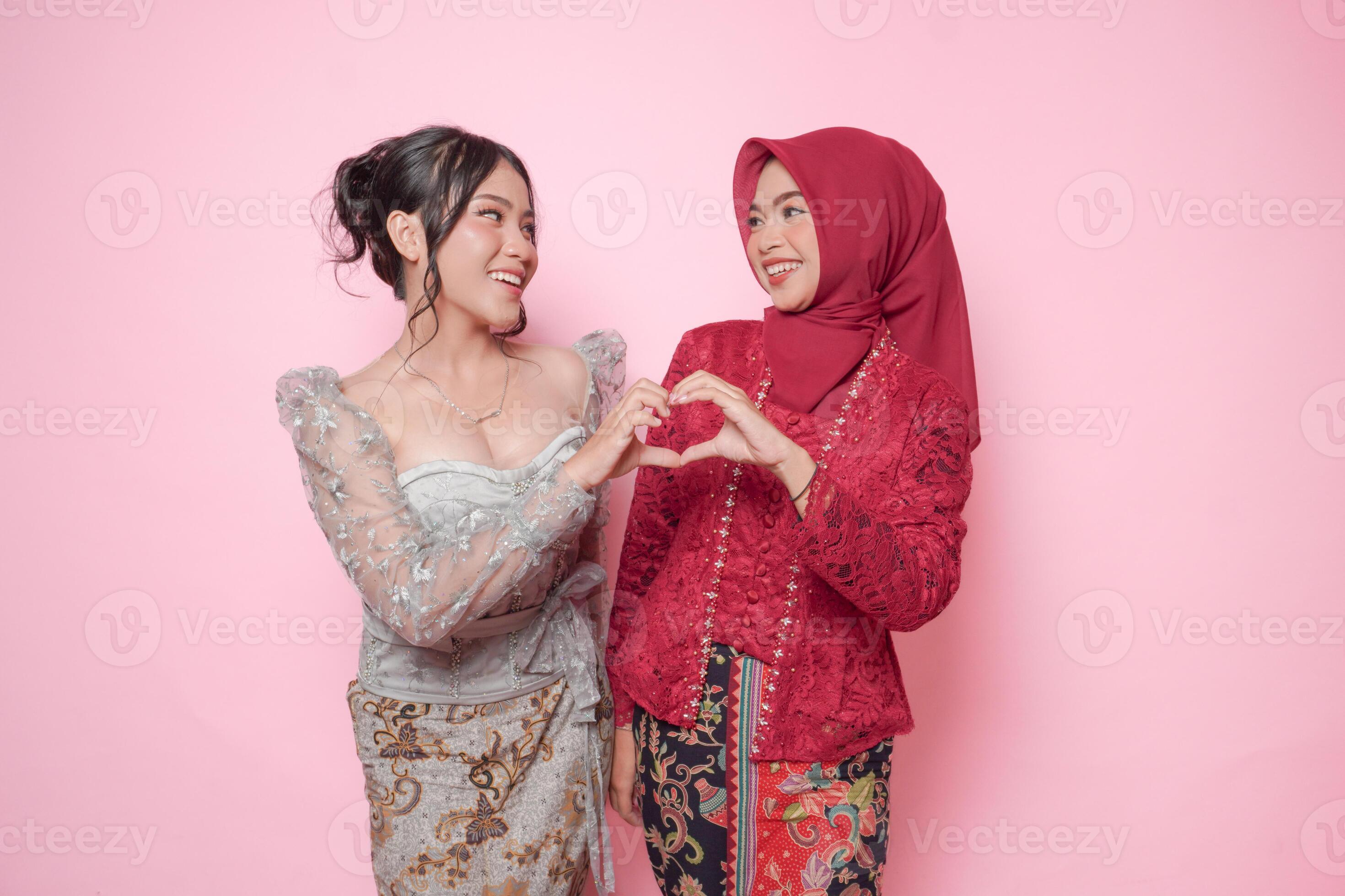 Excited two Asian young woman wearing modern kebaya making heart shape gesture together ...