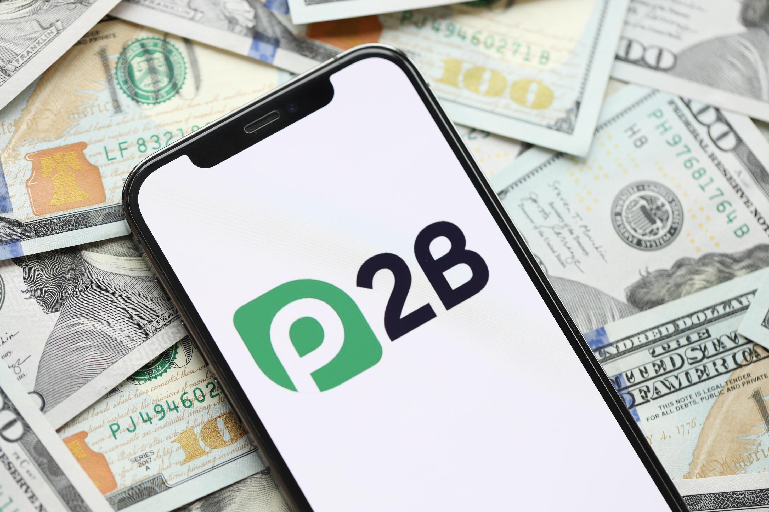KYIV, UKRAINE - MARCH 15, 2024 P2B logo on iPhone display screen with many hundred dollar bills ...