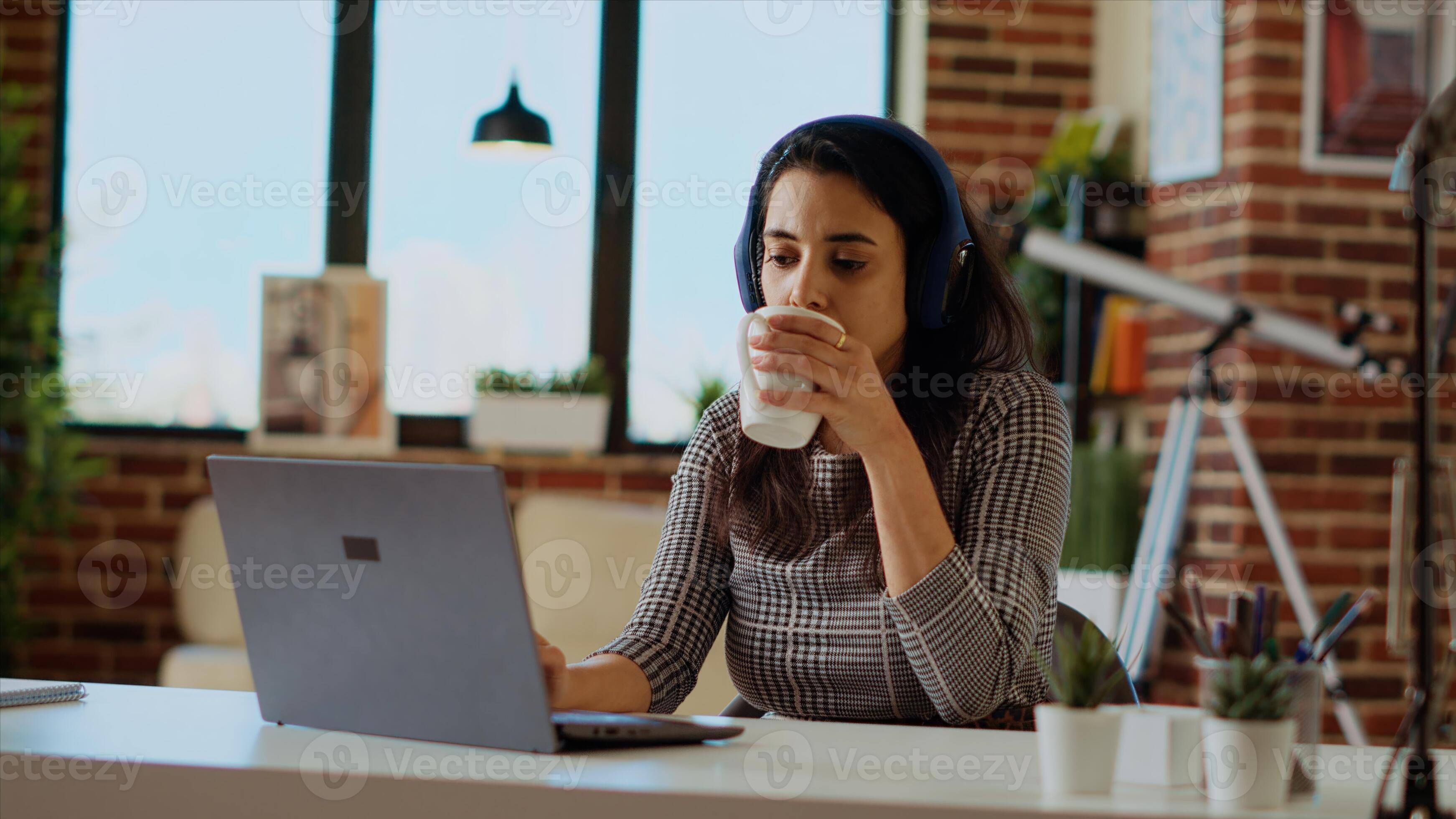 Teleworker checking emails on laptop while at computer desk, drinking coffee and listening music ...