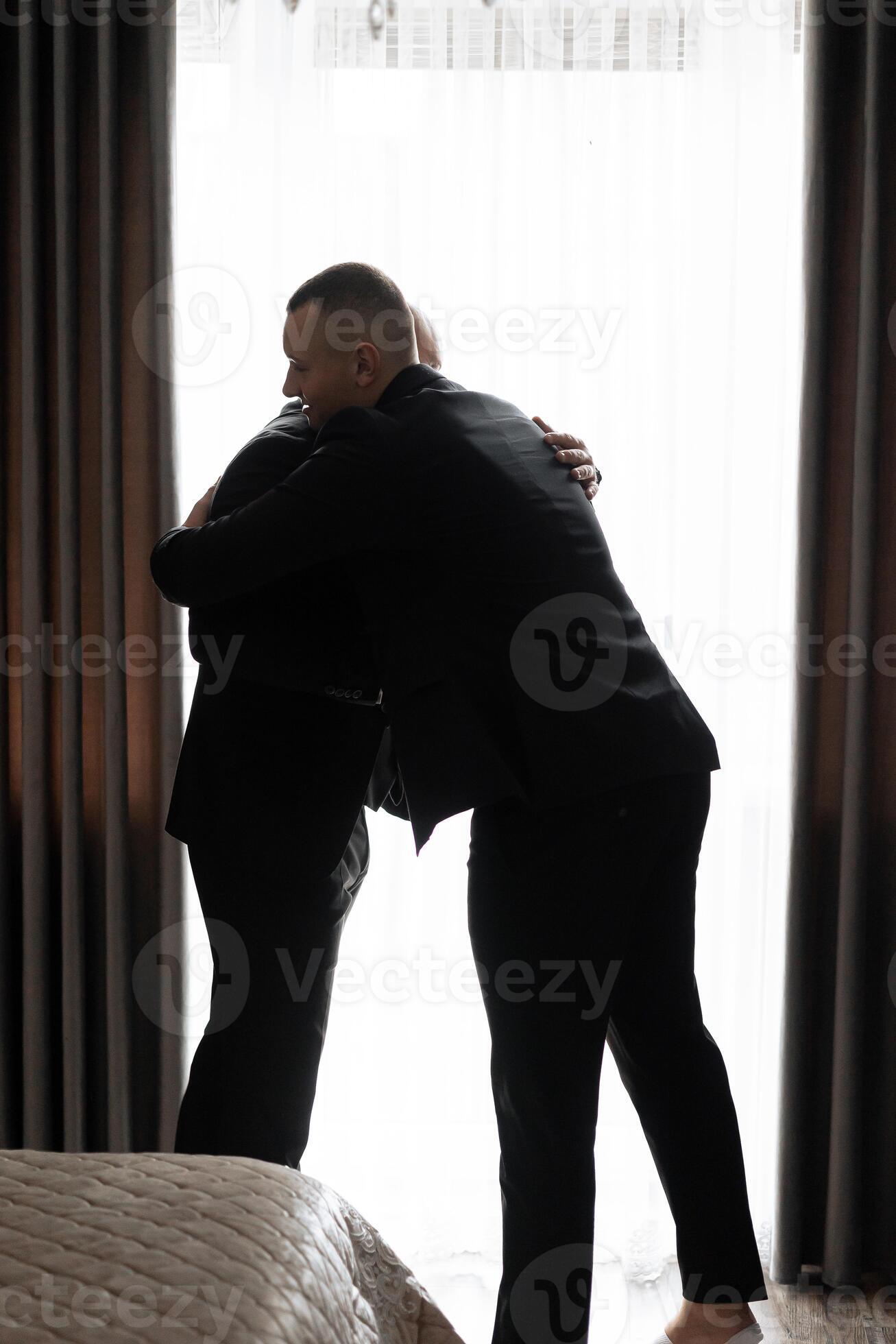 Two men hug each other in a hotel room. One of them is wearing a suit. Scene is warm and ...