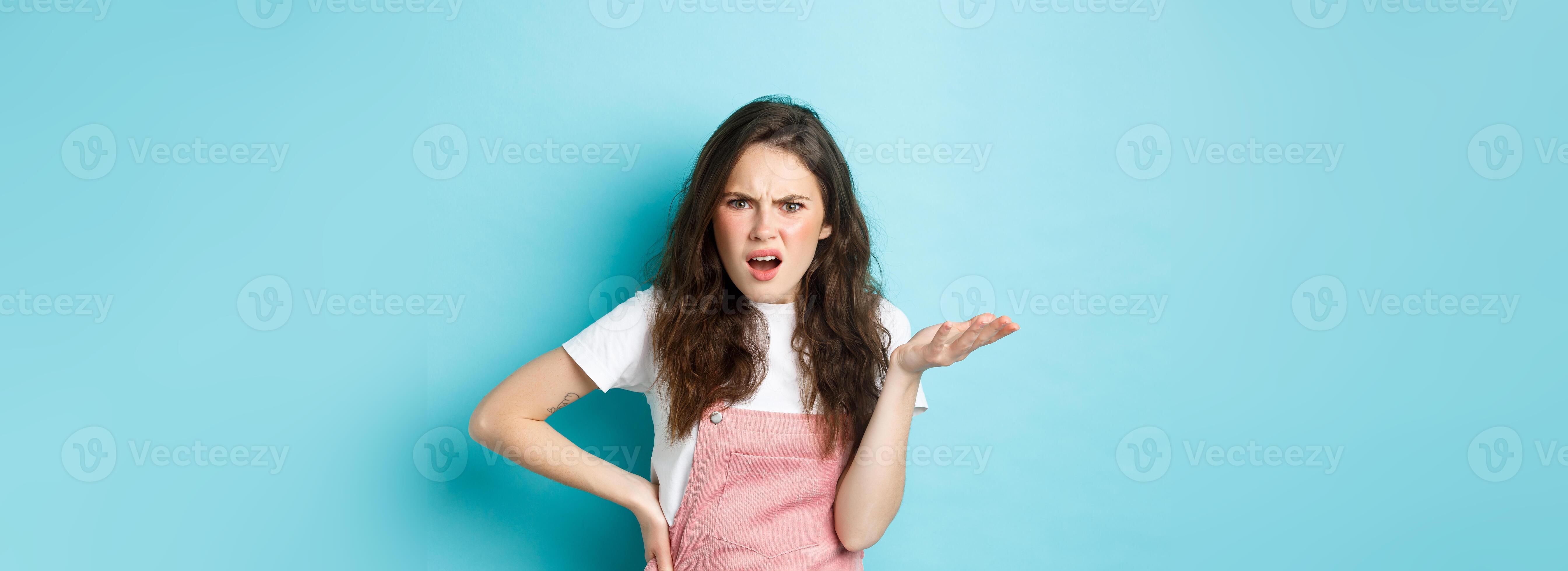 Image of confused and annoyed young woman raising hand and pointing at ...