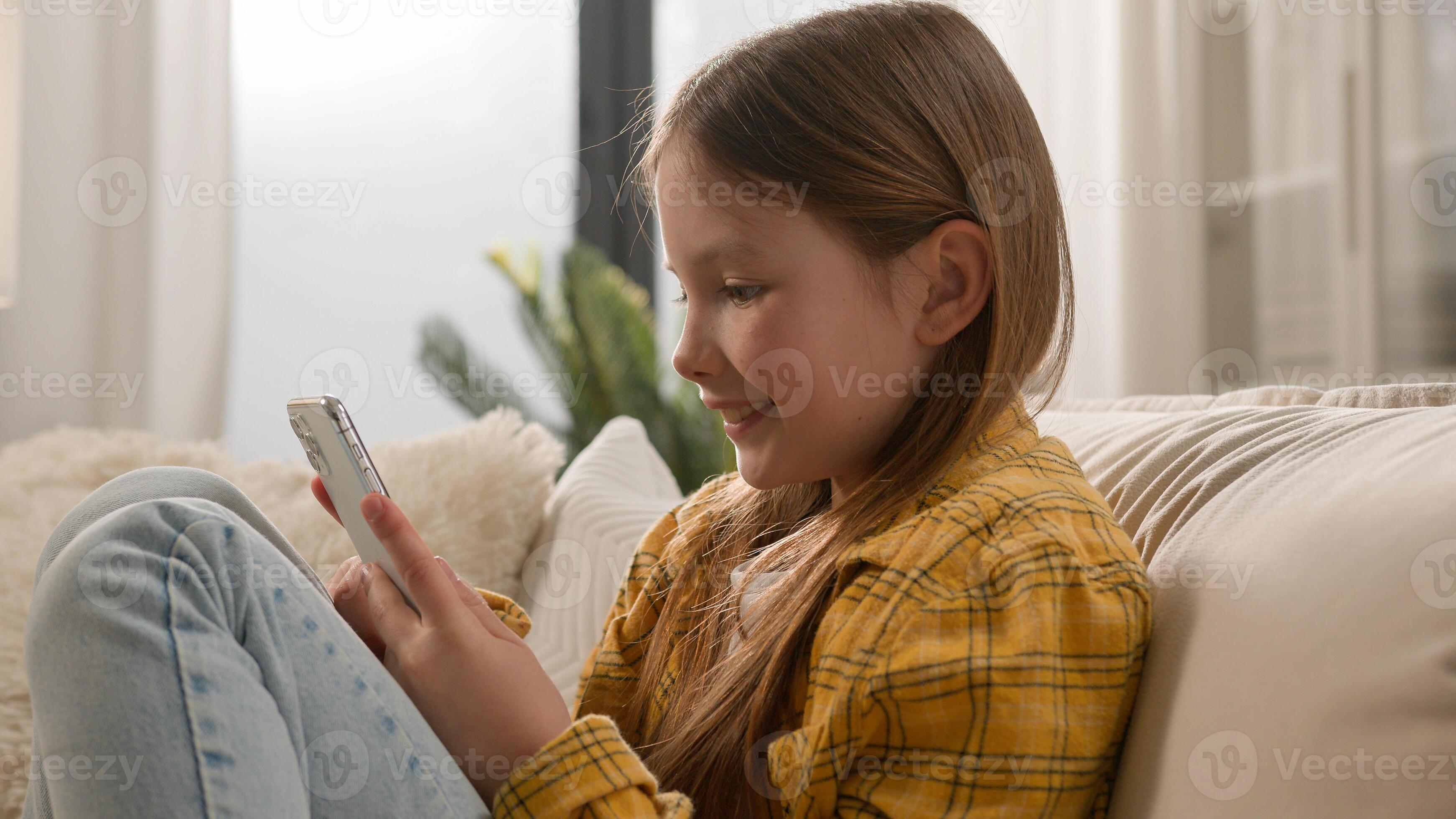 Smiling carefree little Caucasian kid girl scrolling typing screen playing games online using ...