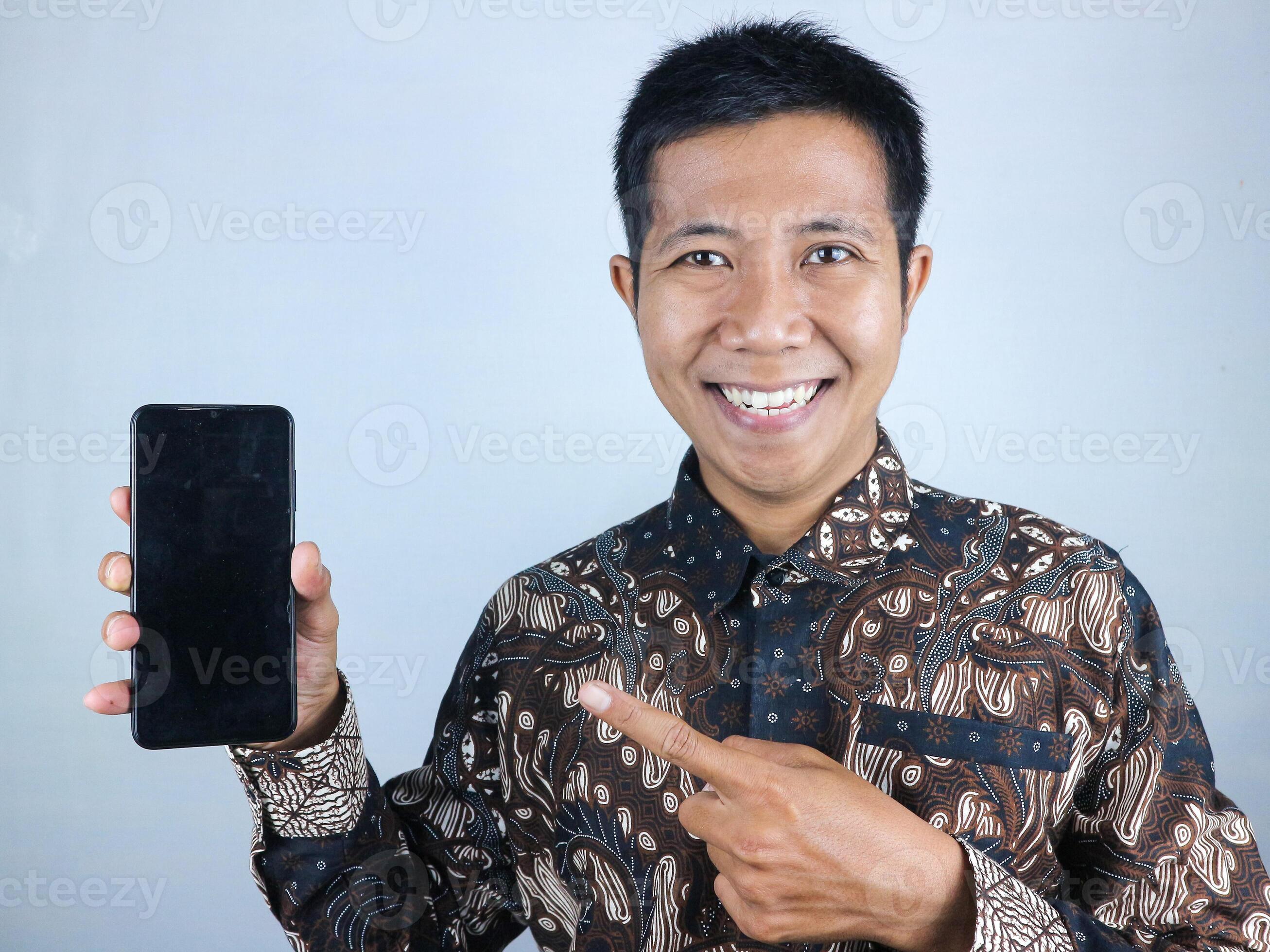 Smiling asian man wearing batik clothes showing smart phone with blank ...