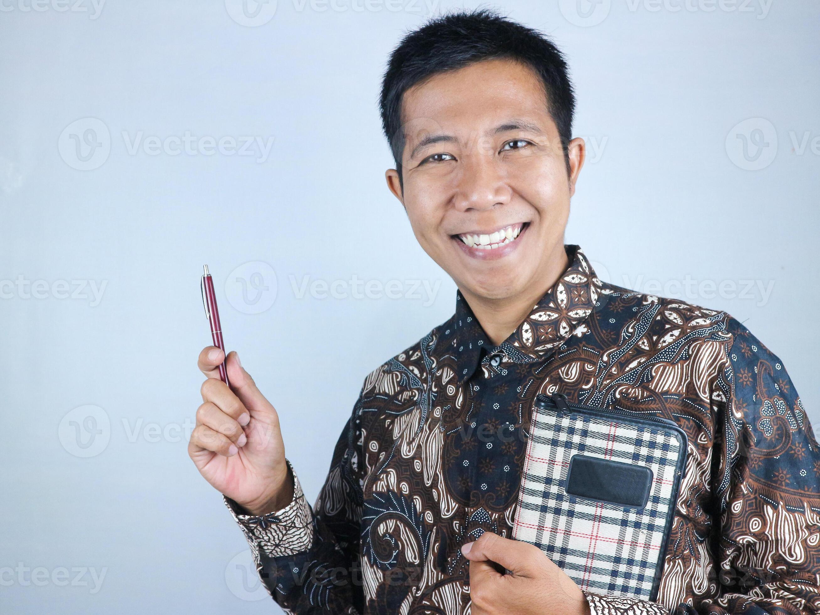 Adult Asian man wearing batik clothes smiling while holding a book and ...