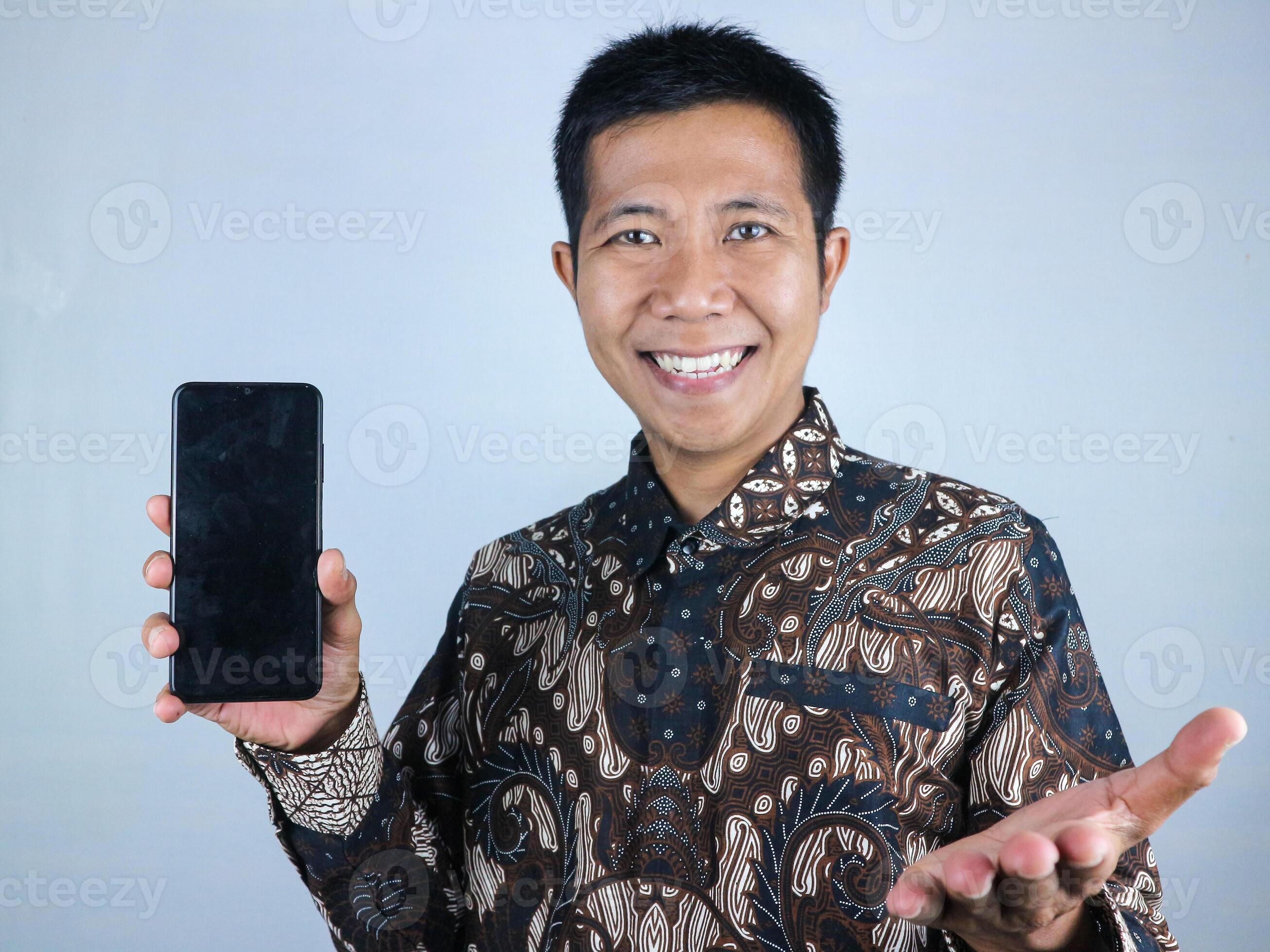 Smiling asian man wearing batik clothes showing smart phone with blank ...