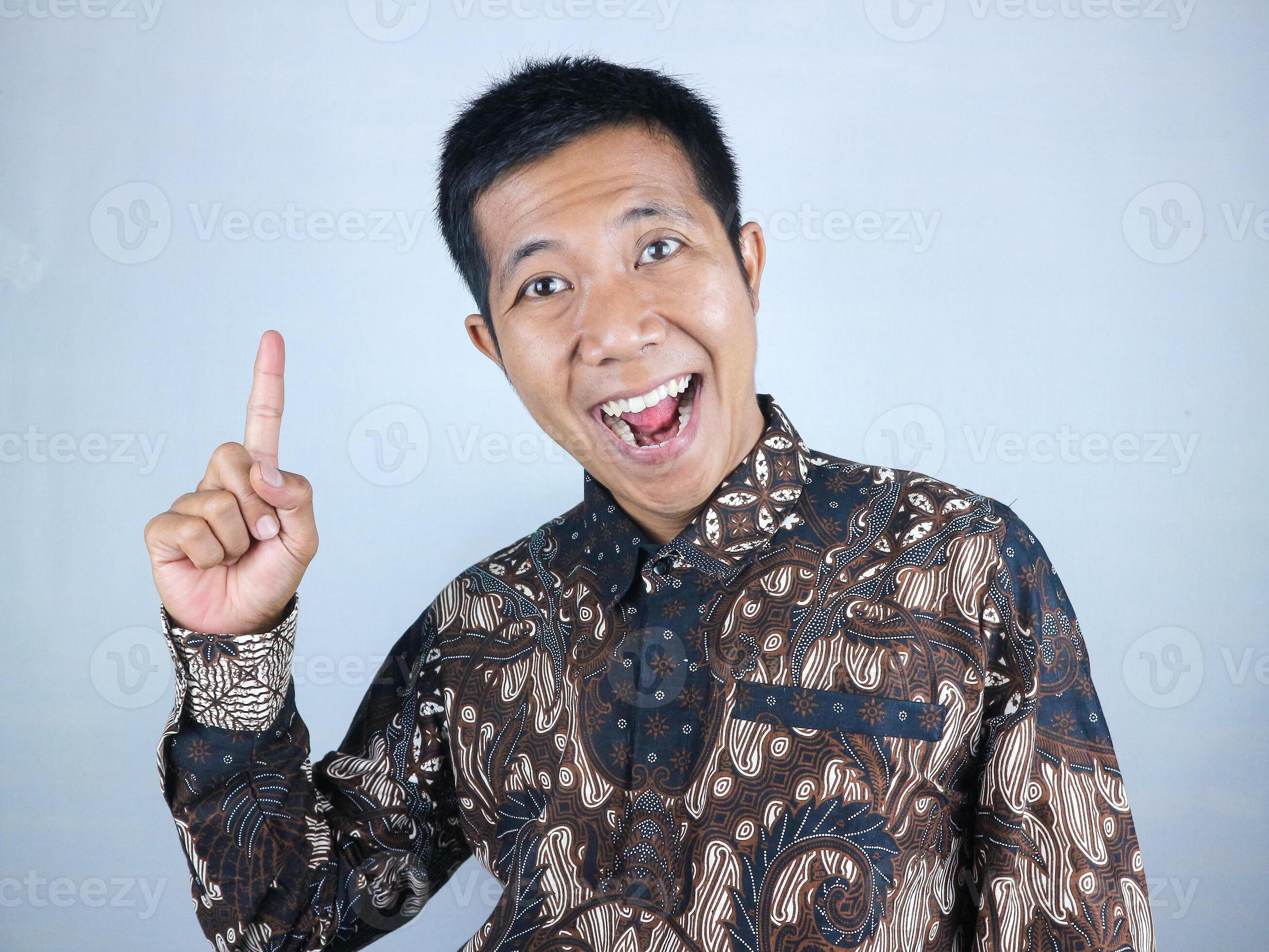 Smiling expression of asian man wearing batik clothes pointing up ...