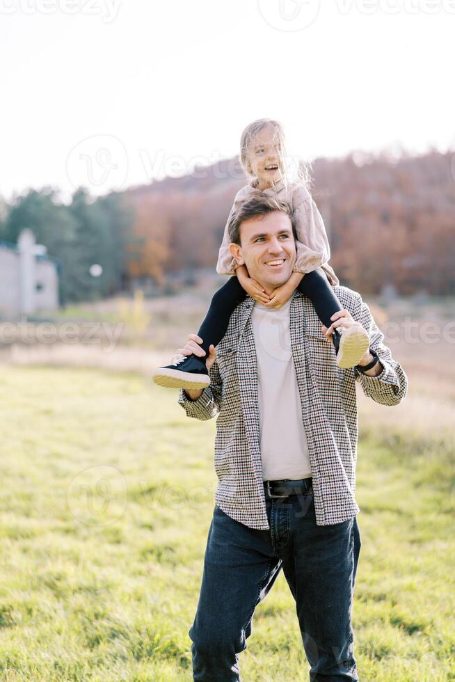 Smiling dad carries laughing little girl on his shoulders across the meadow, looking to the side ...