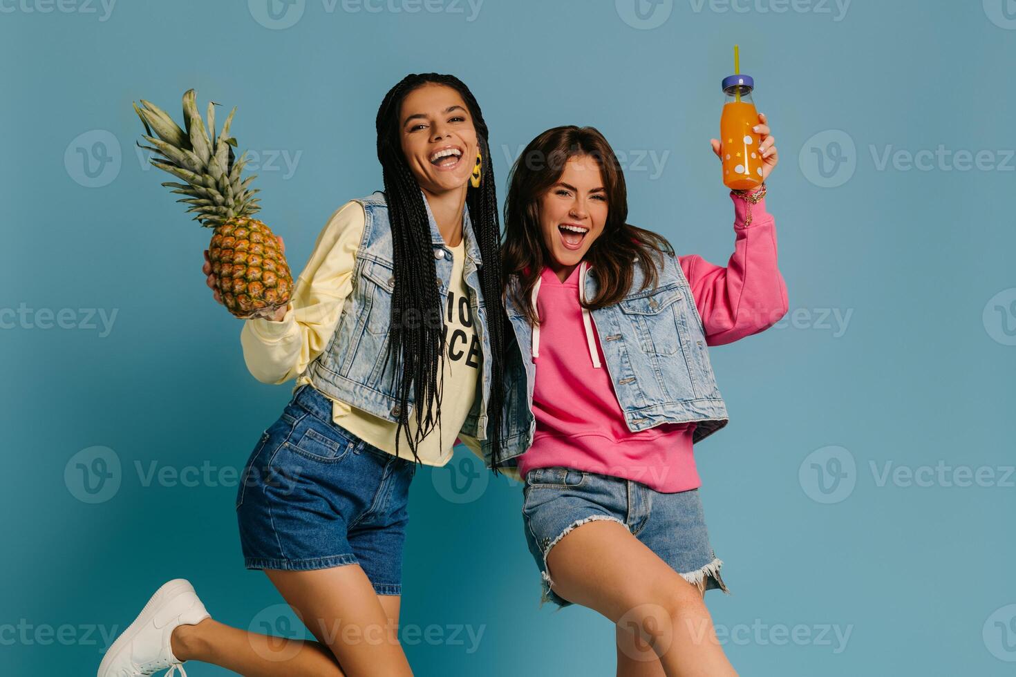 Two playful young women holding pineapple and bottle with lemonade against blue background ...
