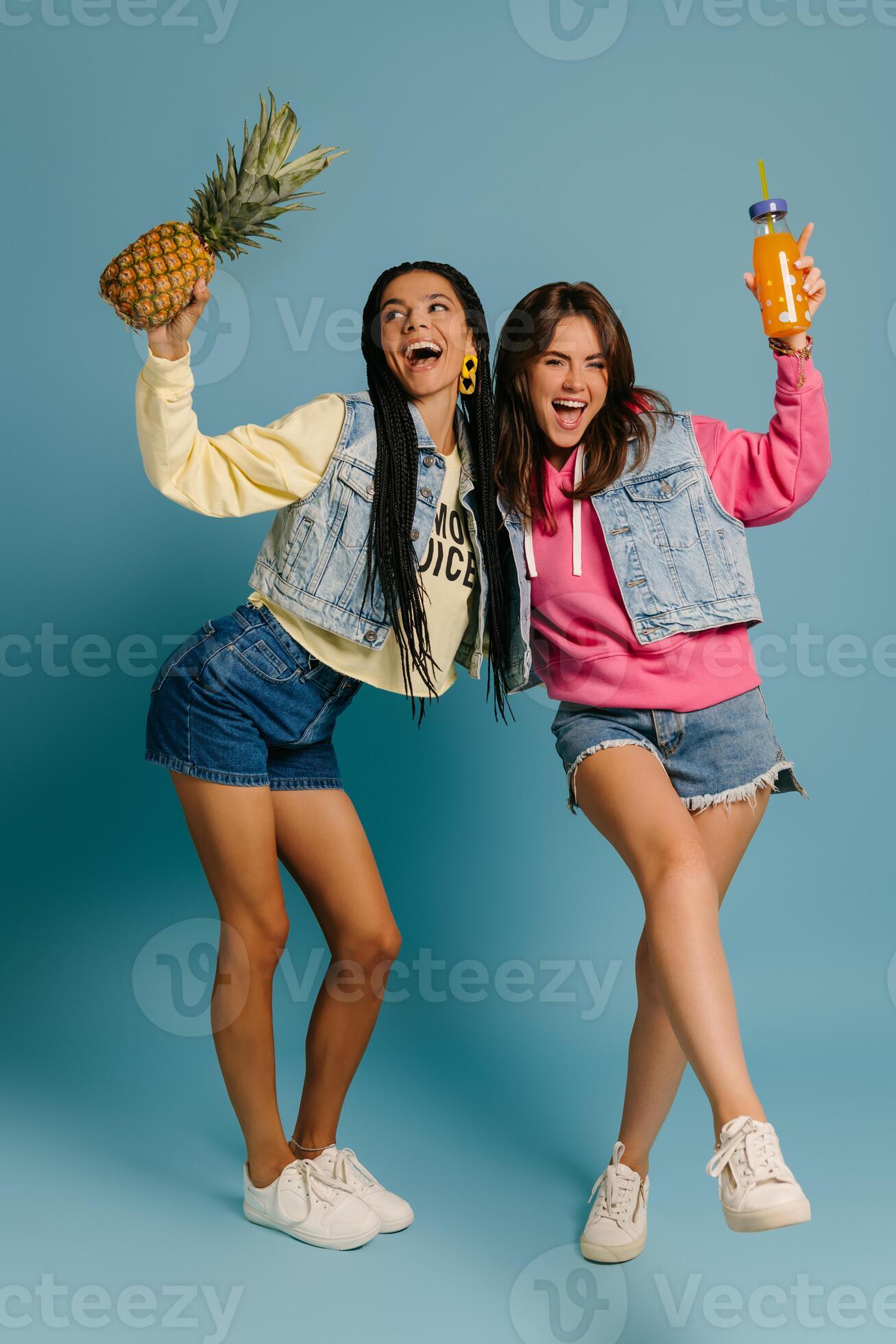 Full length of two playful young women holding pineapple and bottle with lemonade against blue ...