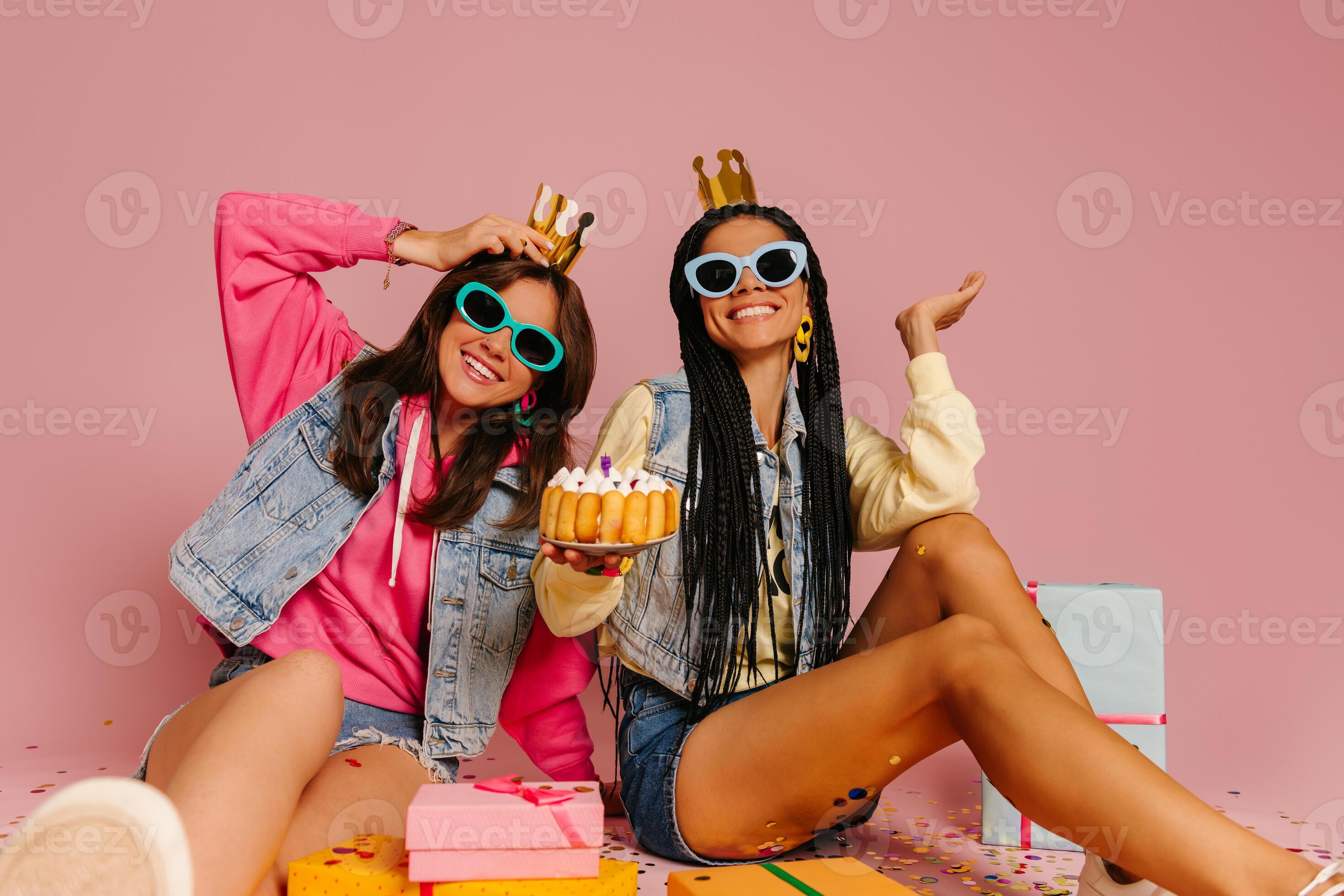 Two playful young women holding birthday cake and smiling while sitting near gift boxes on pink ...