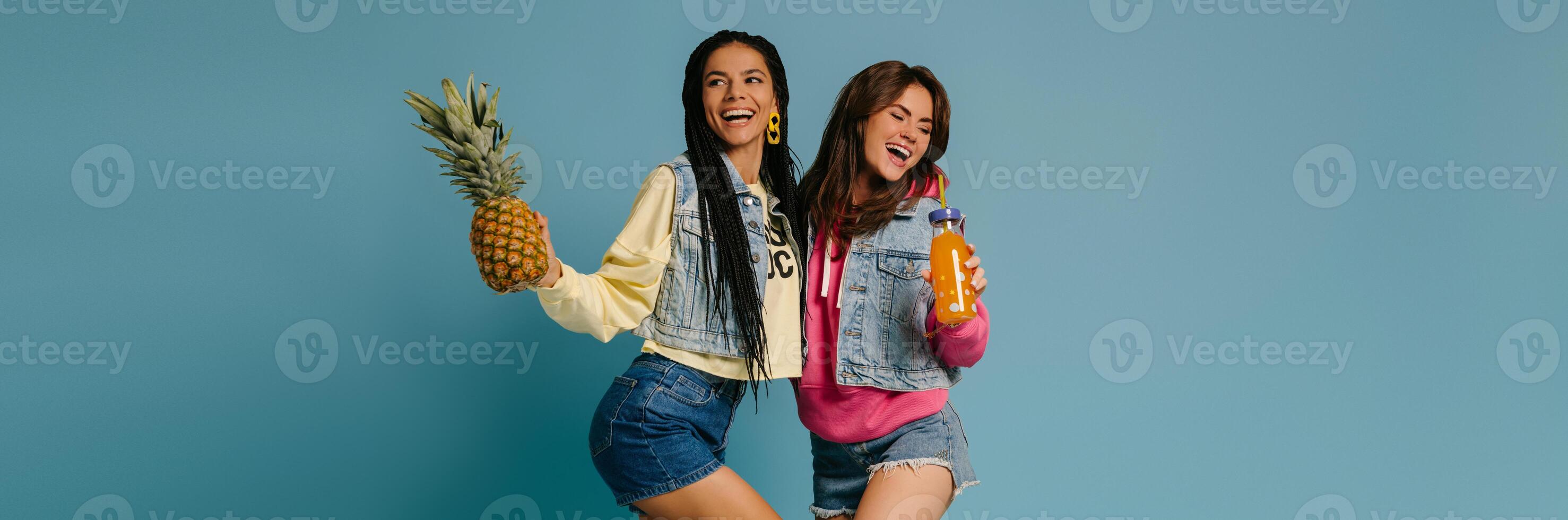 Two playful young women holding pineapple and bottle with lemonade against blue background ...