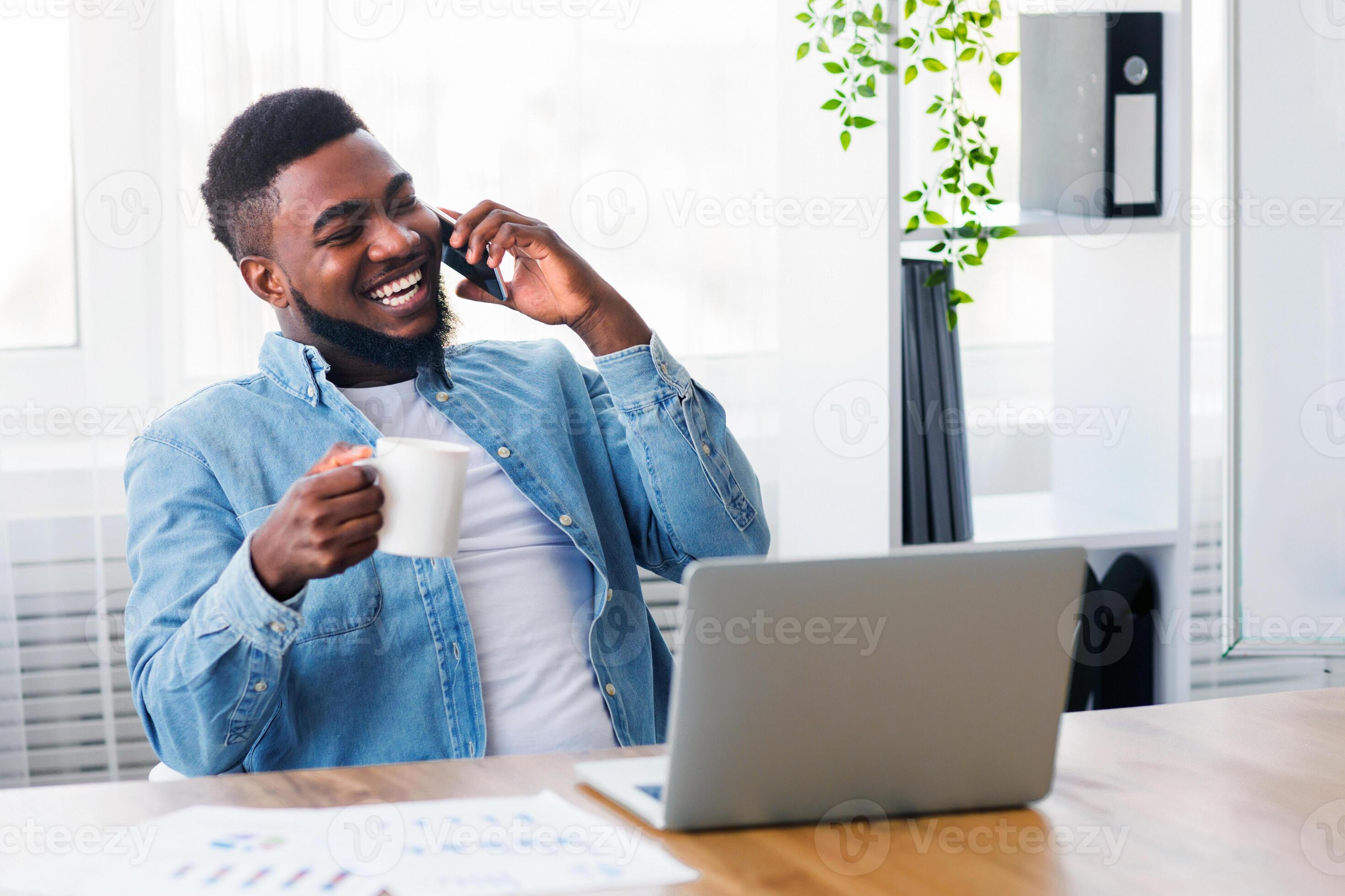 Cheerful black office worker talking on phone, laughing and drinking coffee at workplace, copy ...