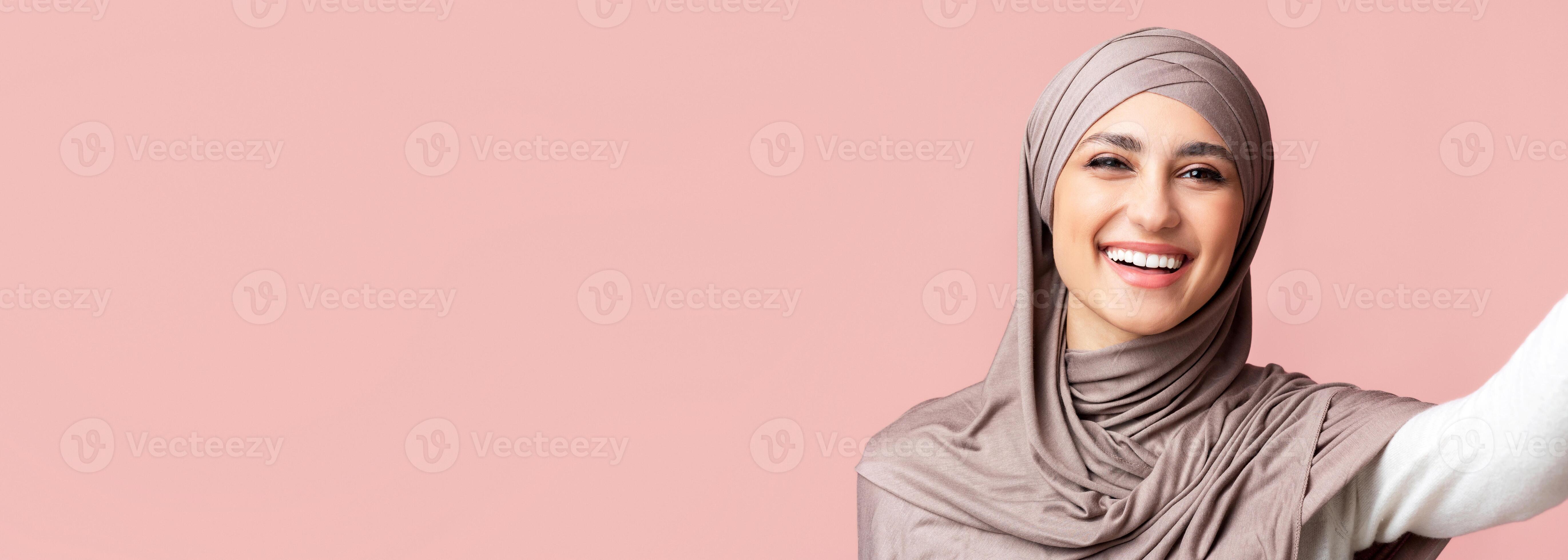 Cheerful arabic girl in hijab taking selfie and sincerely laughing, pink background with free ...