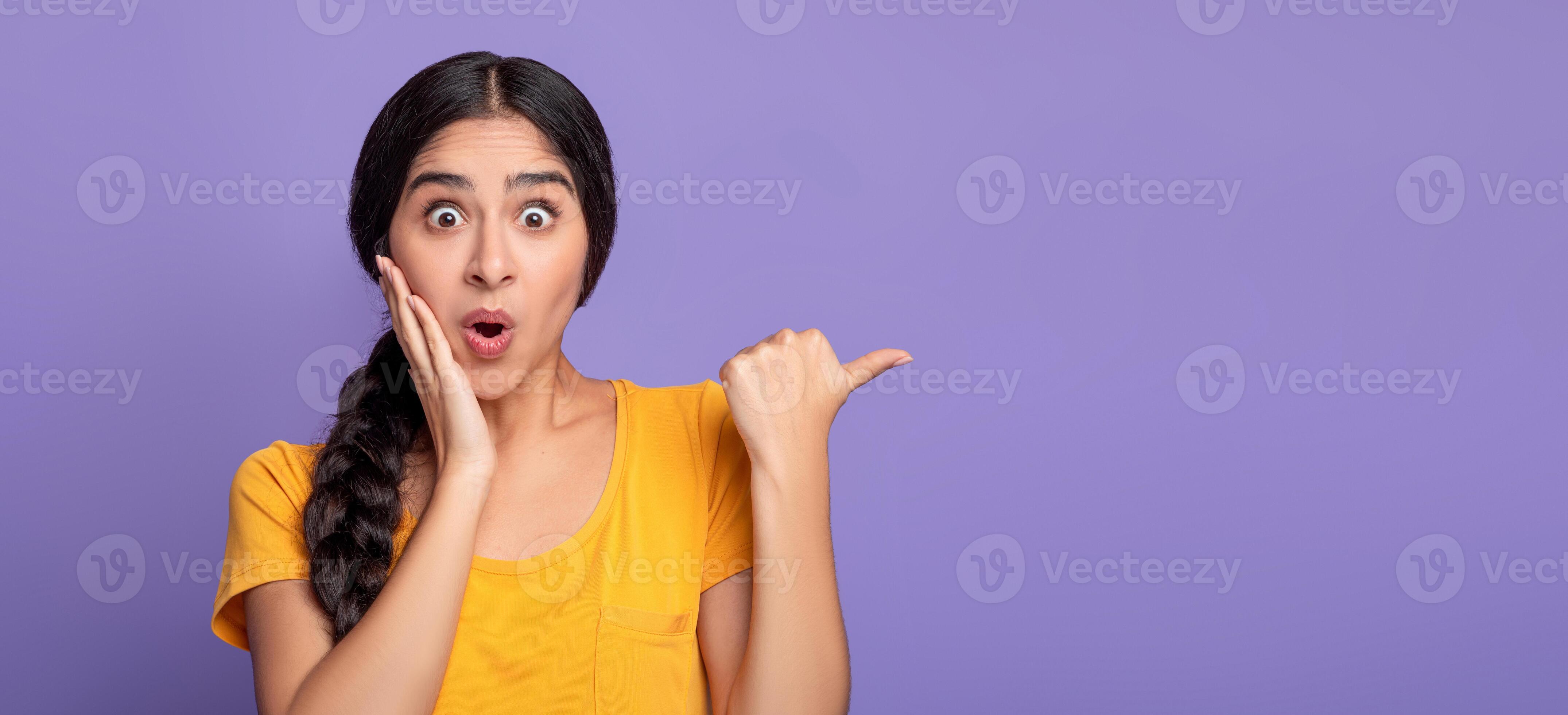 Omg, Cool Offer. Surprised young indian lady pointing at free space isolated over purple studio ...