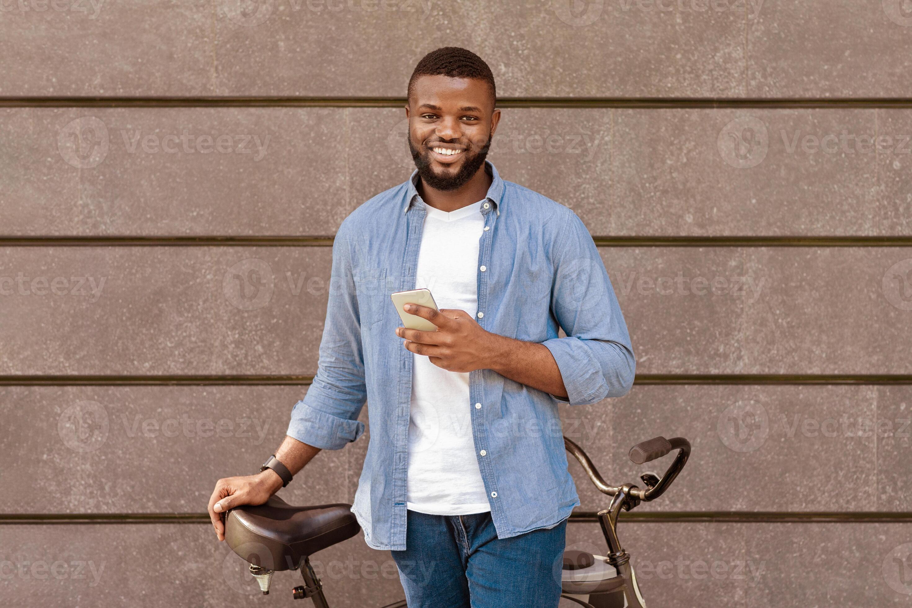 Outdoor portrait of handsome afro guy with smartphone and bike posing over gray urban wall ...