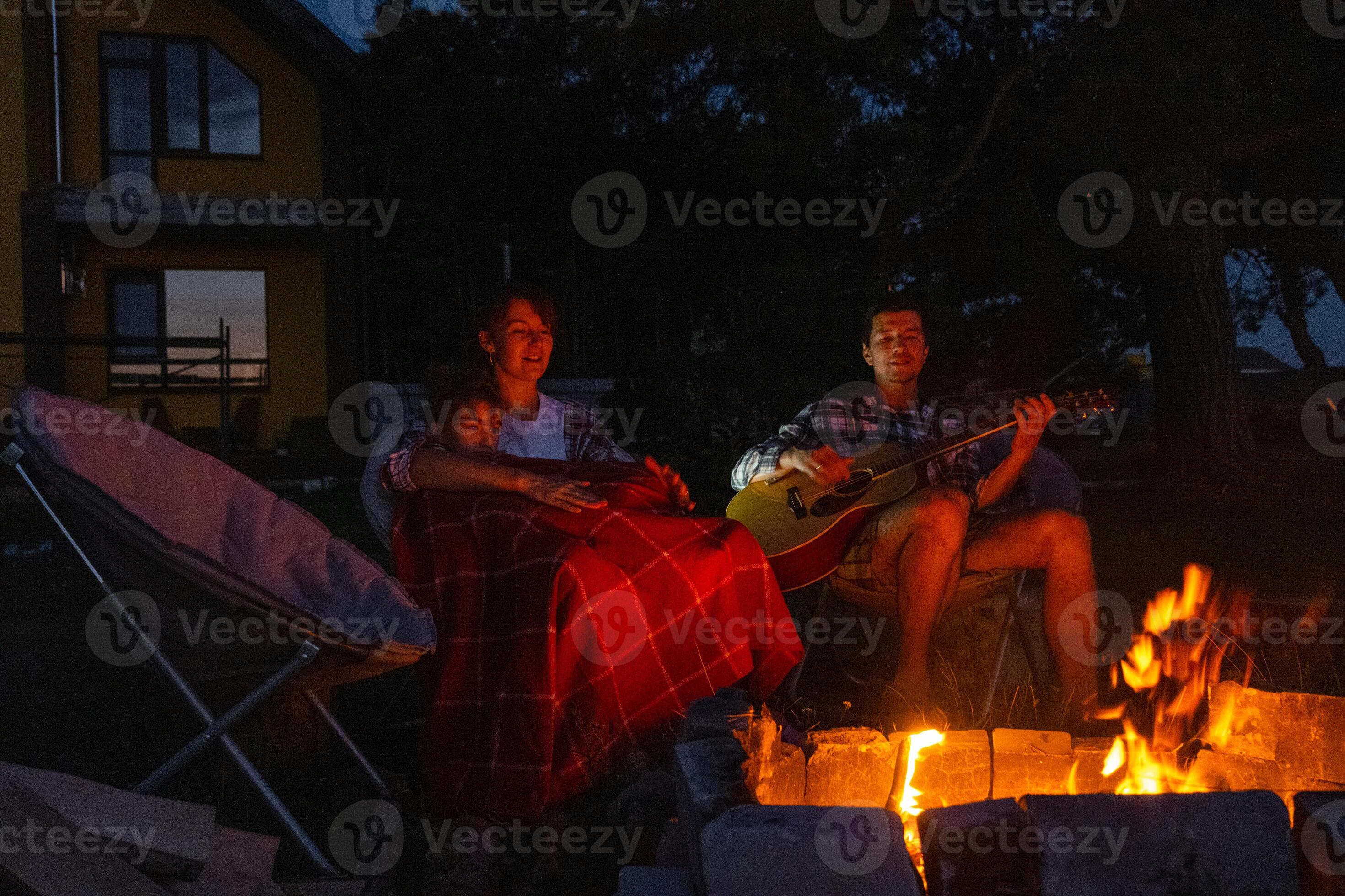 The family is sitting around the campfire, man plays the guitar, a woman listens and sings along ...