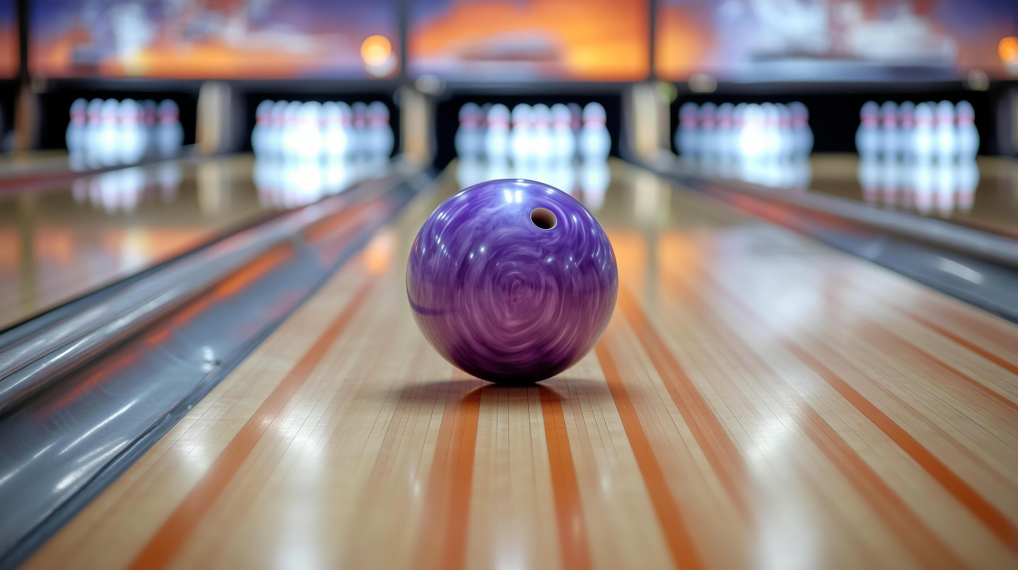 Purple bowling ball rolling down lane with pins in background at bowling alley during evening ...