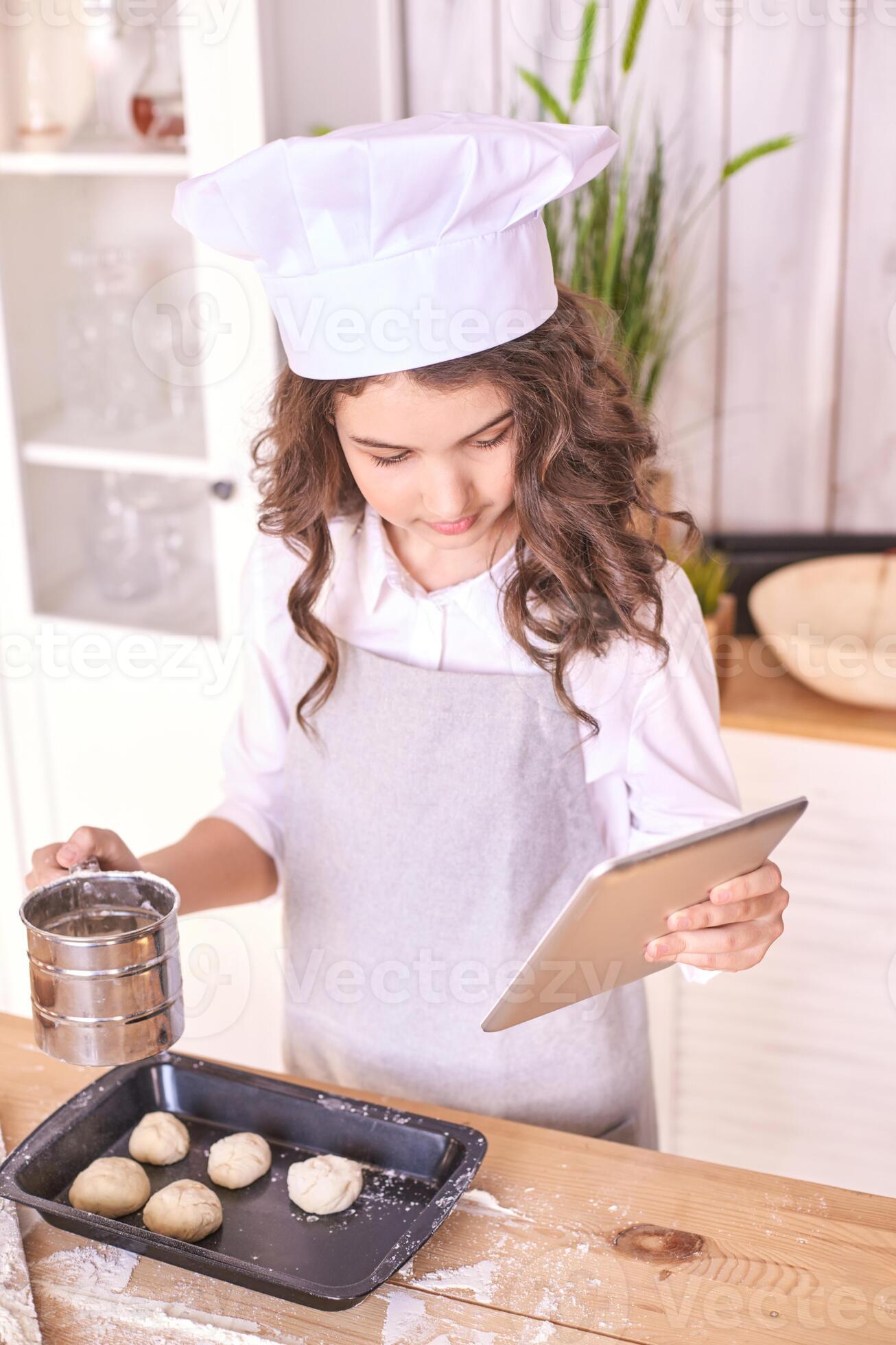 Young girl chef sifting flour on dough in kitchen while learning recipe from tablet for baking ...