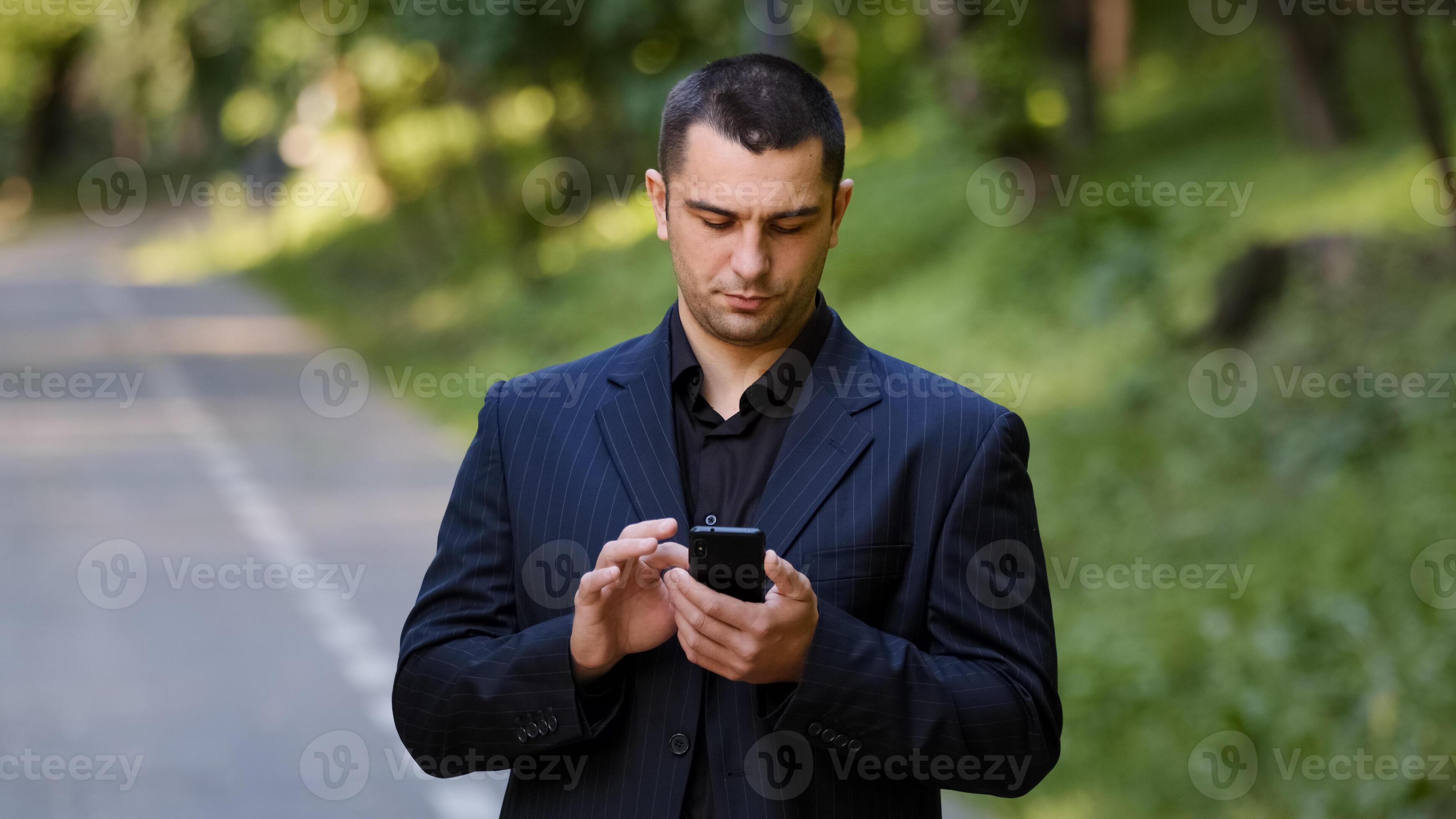 Serious adult caucasian man user stand outdoors in park wears black formal suit hold mobile ...